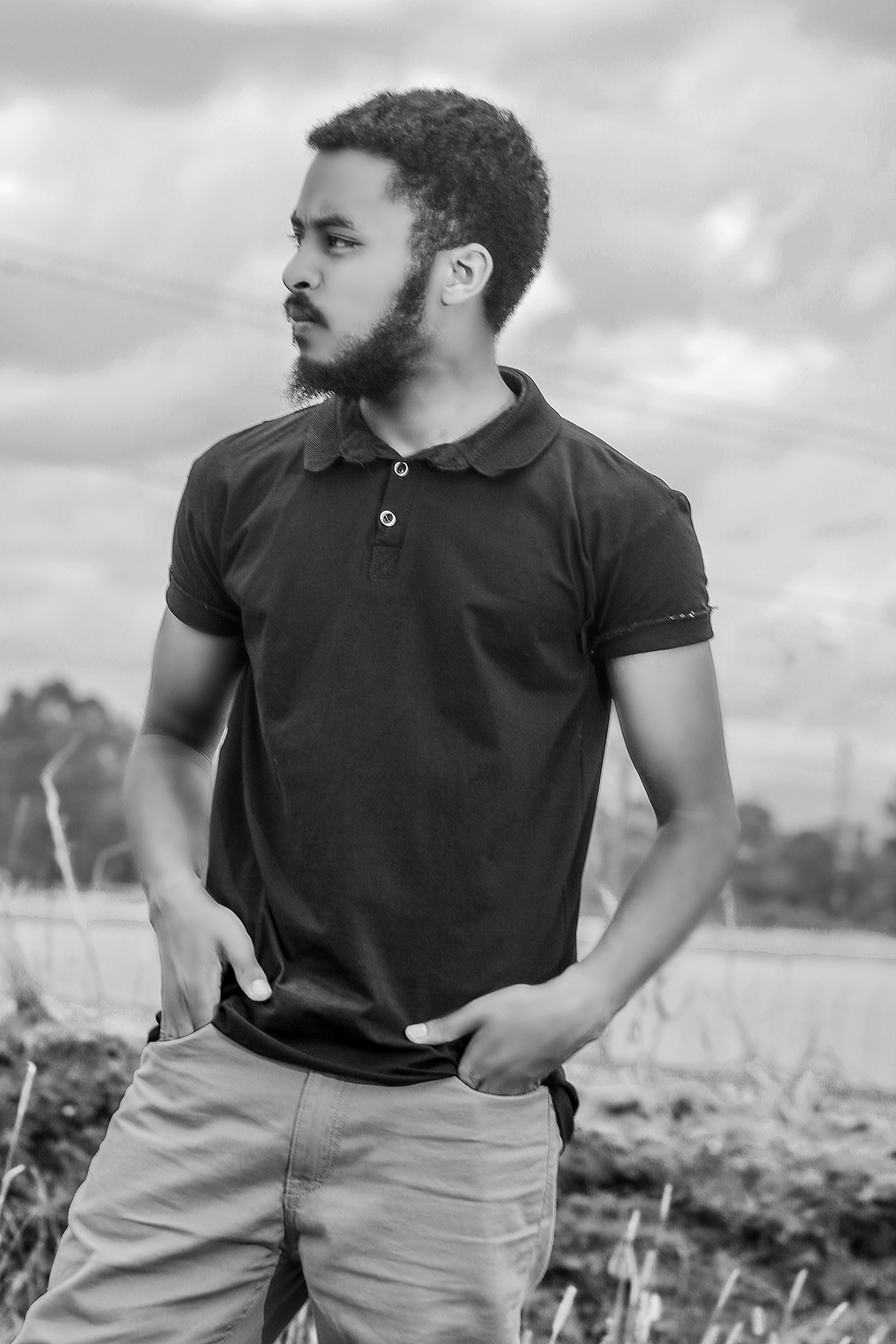 A young man in a black polo shirt stands thoughtfully outdoors, with his hands in his pockets and a moody sky in the background.