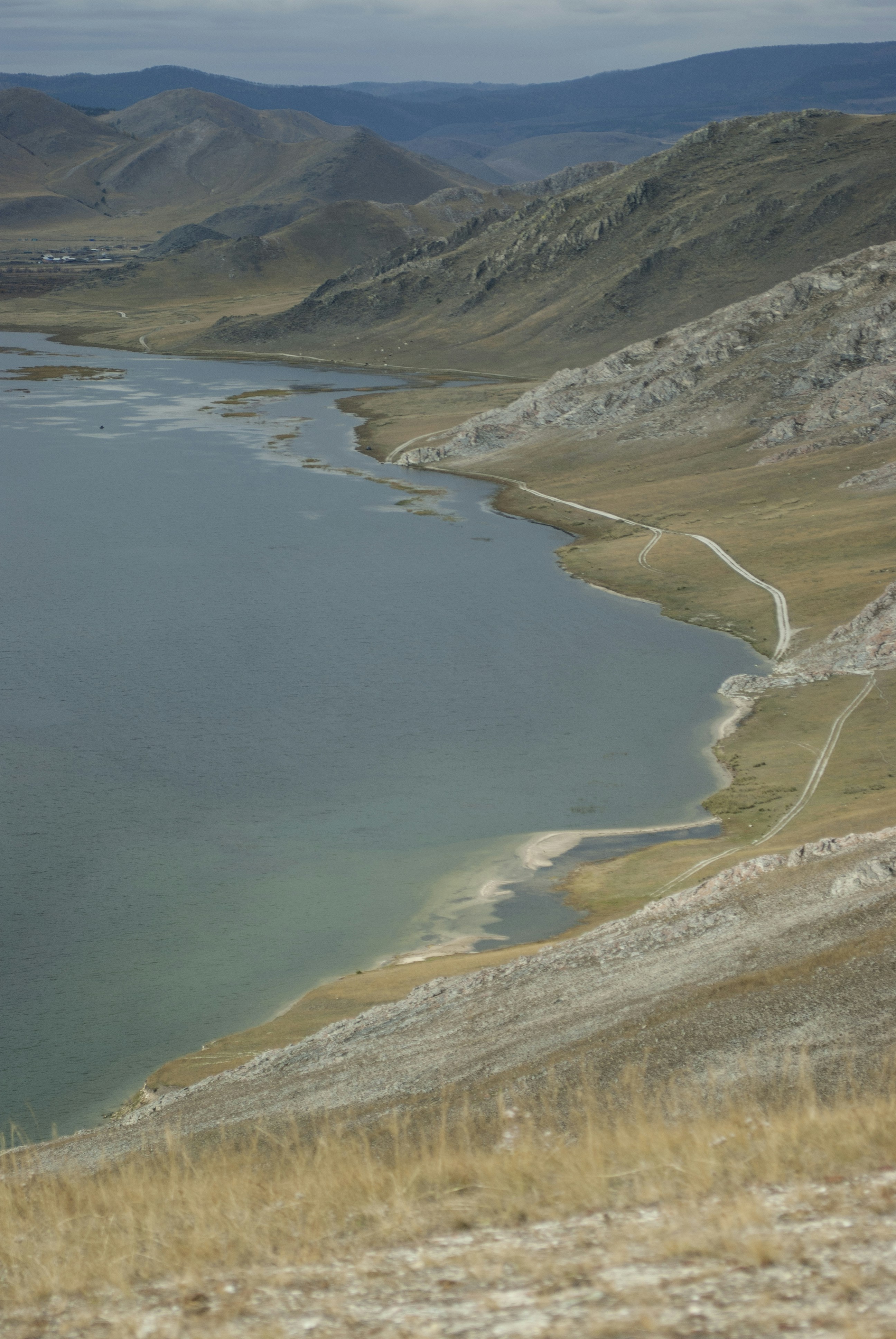Coastal landscape with a winding road along the hillside beside a calm bay under a cloudy sky.
