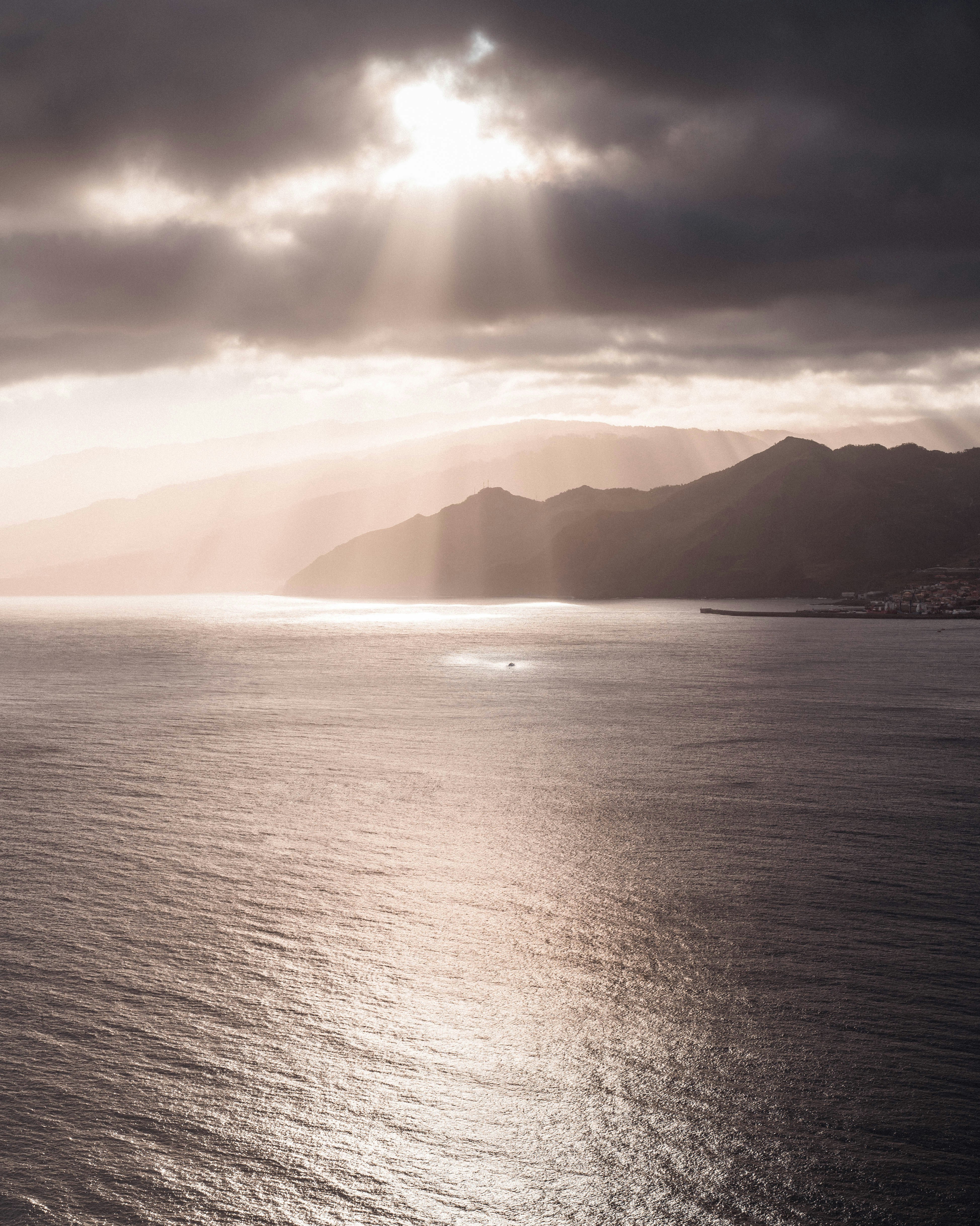 Sun rays break through heavy clouds onto a calm fjord, creating a luminous path along the water toward distant mountains.