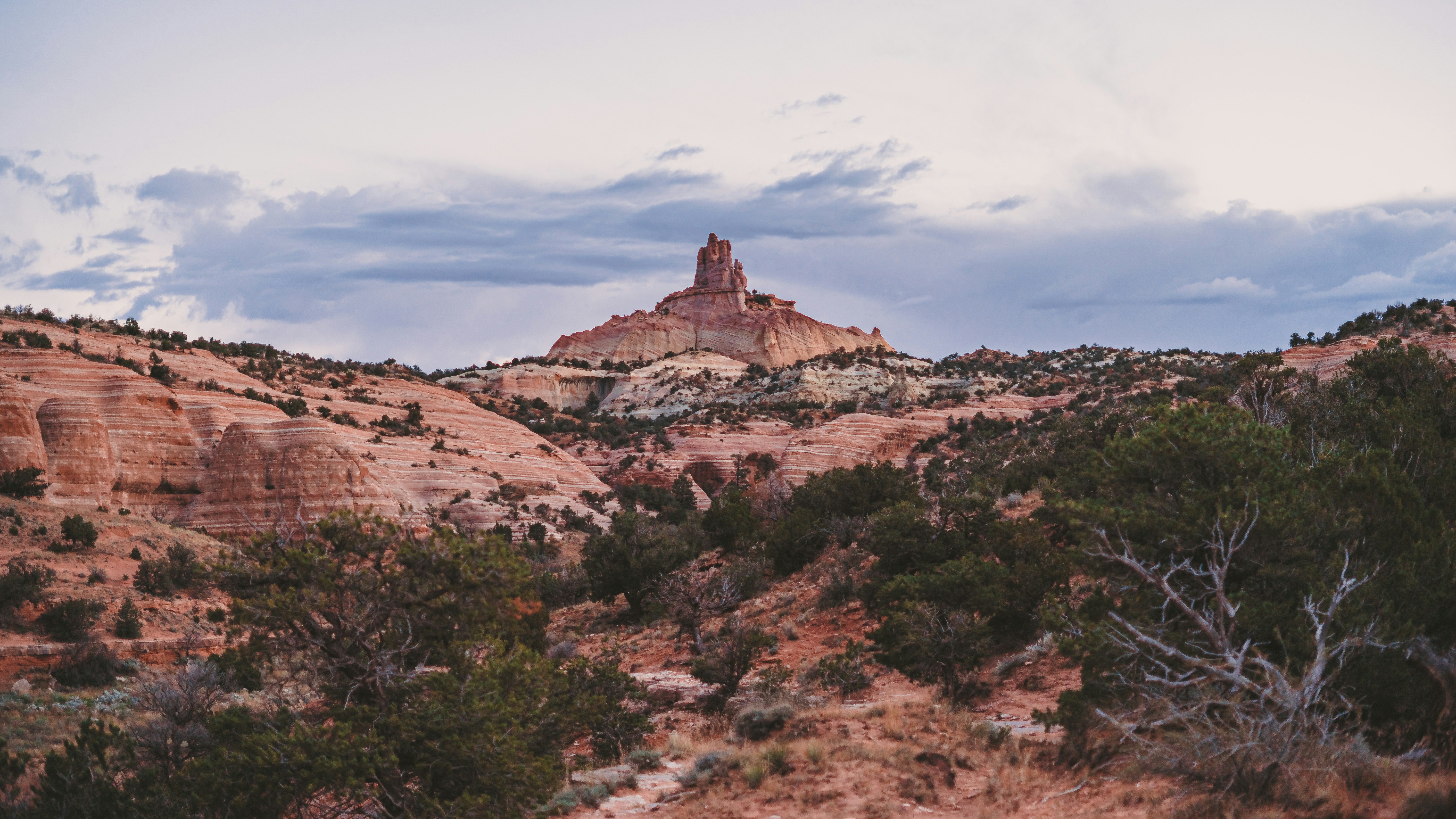 A majestic rock formation towers over the arid landscape, surrounded by rugged terrain and sparse vegetation during twilight.