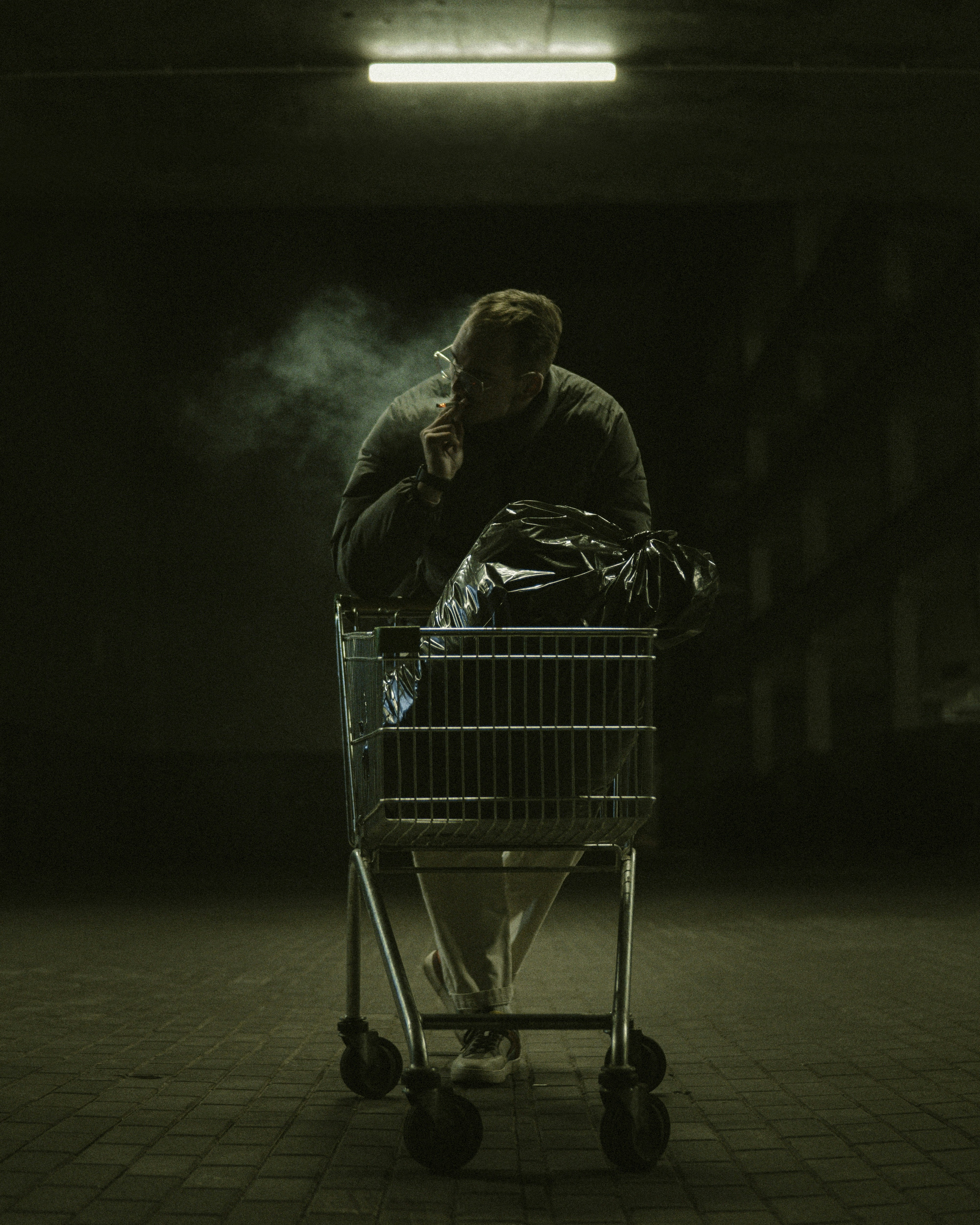 A man stands by a shopping cart filled with a trash bag, exhaling smoke in a dimly lit parking garage.