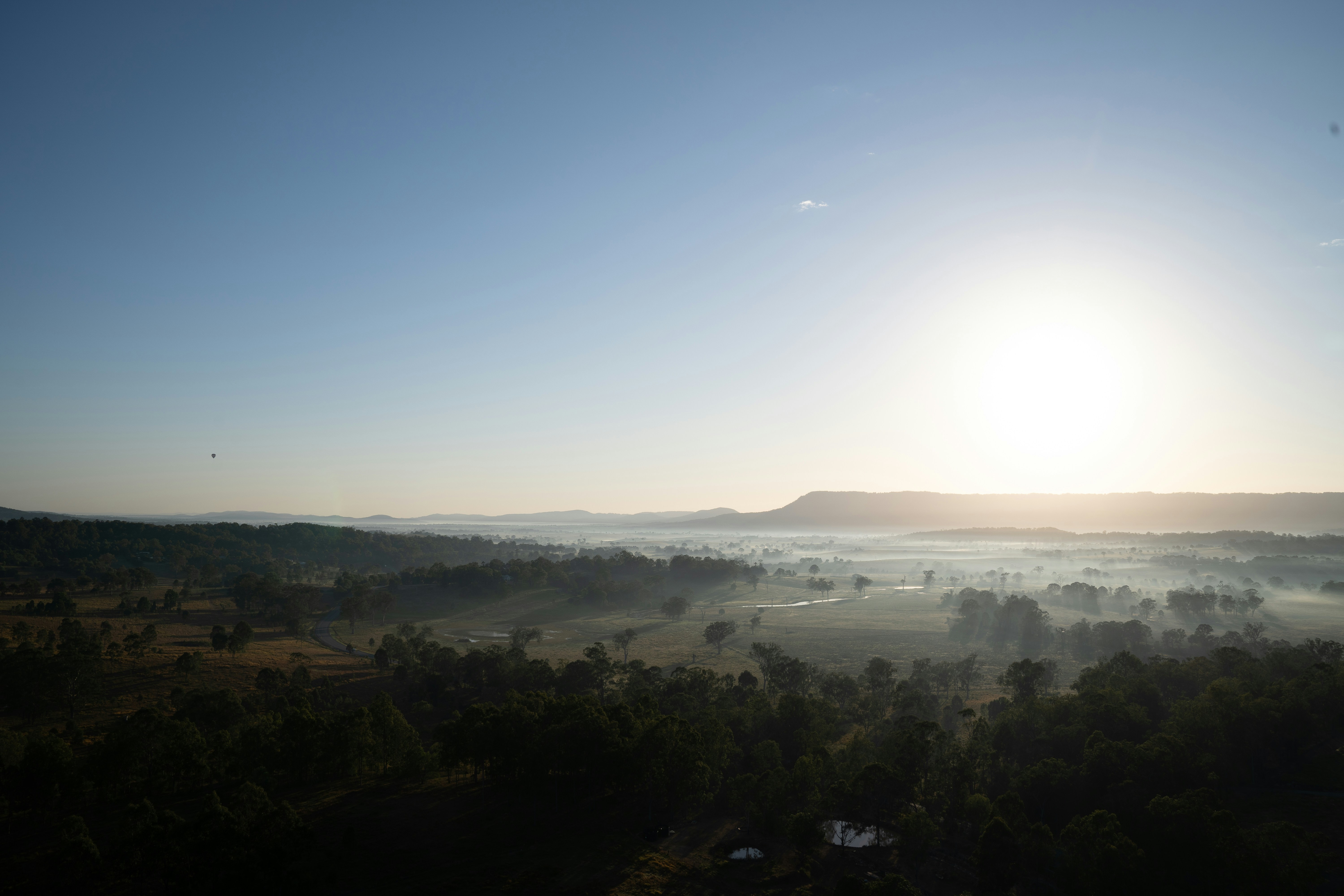 A panoramic view of a tranquil valley shrouded in morning mist, with the sun rising over distant hills. The landscape showcases lush greenery and soft light.