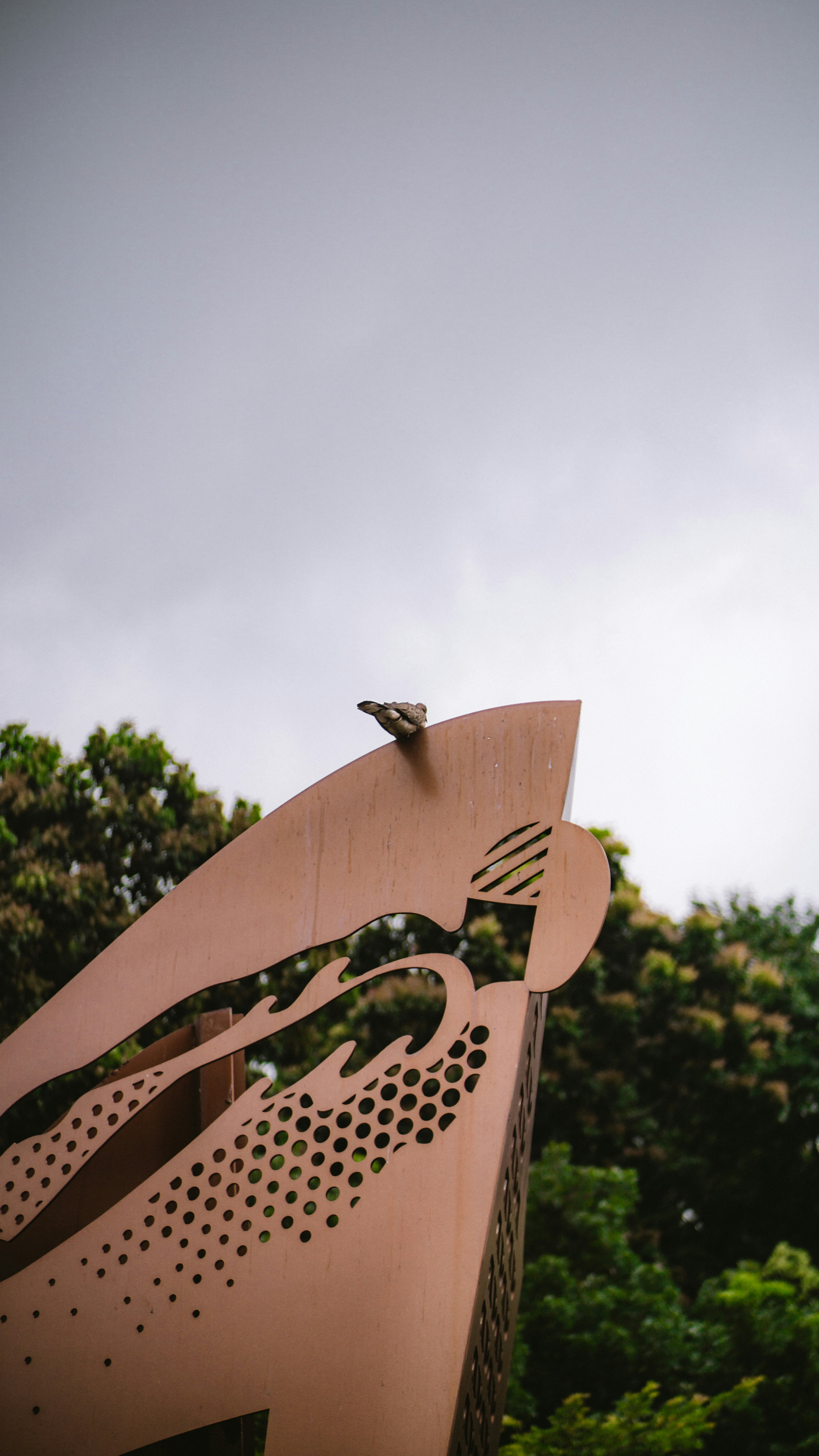 Perched on Rust: A Bird atop a Perforated Metal Monument against a Cloudy Sky
