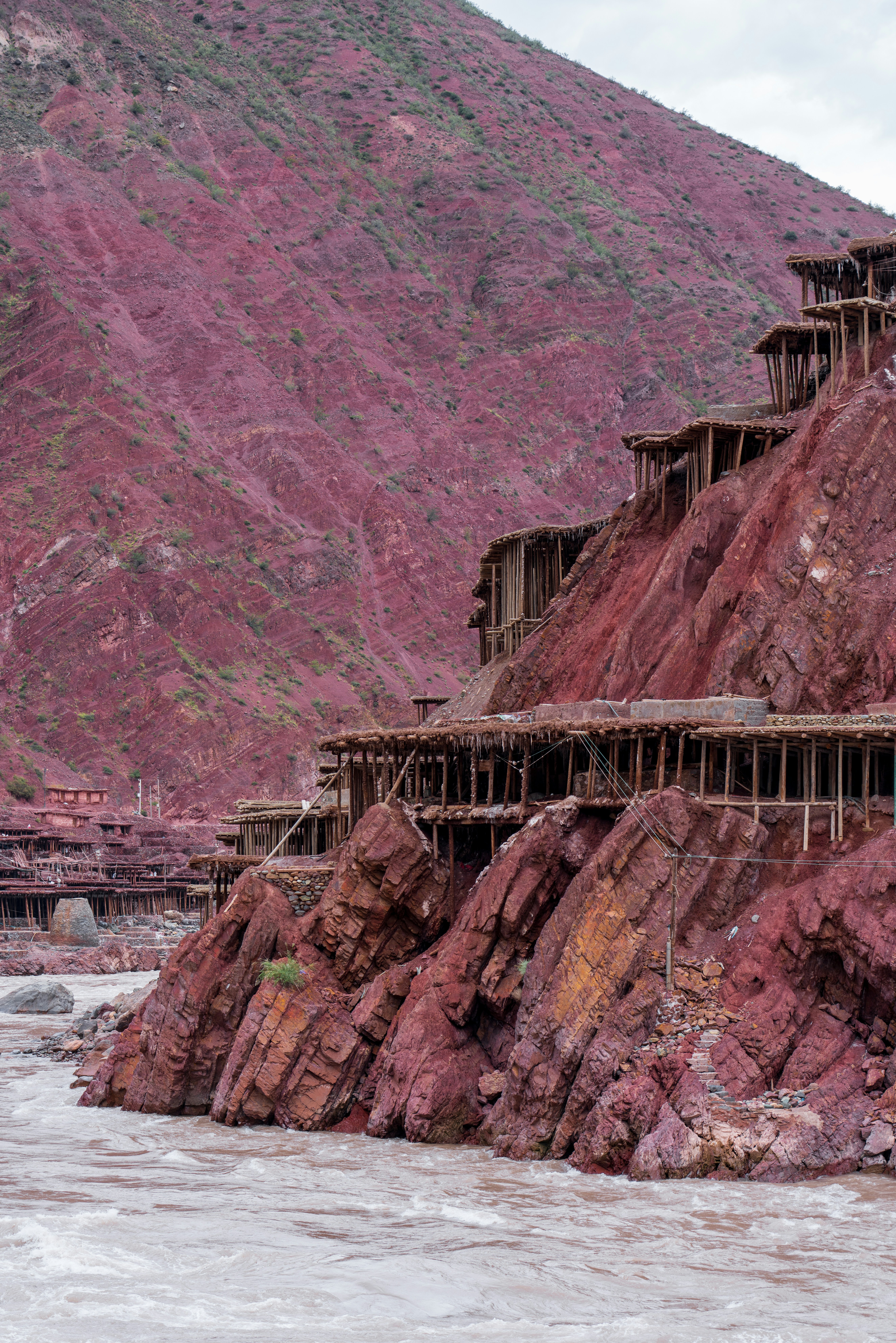 Ancient wooden structures cling to the steep red cliffs, reflecting a bygone era of industriousness against the rushing river below.