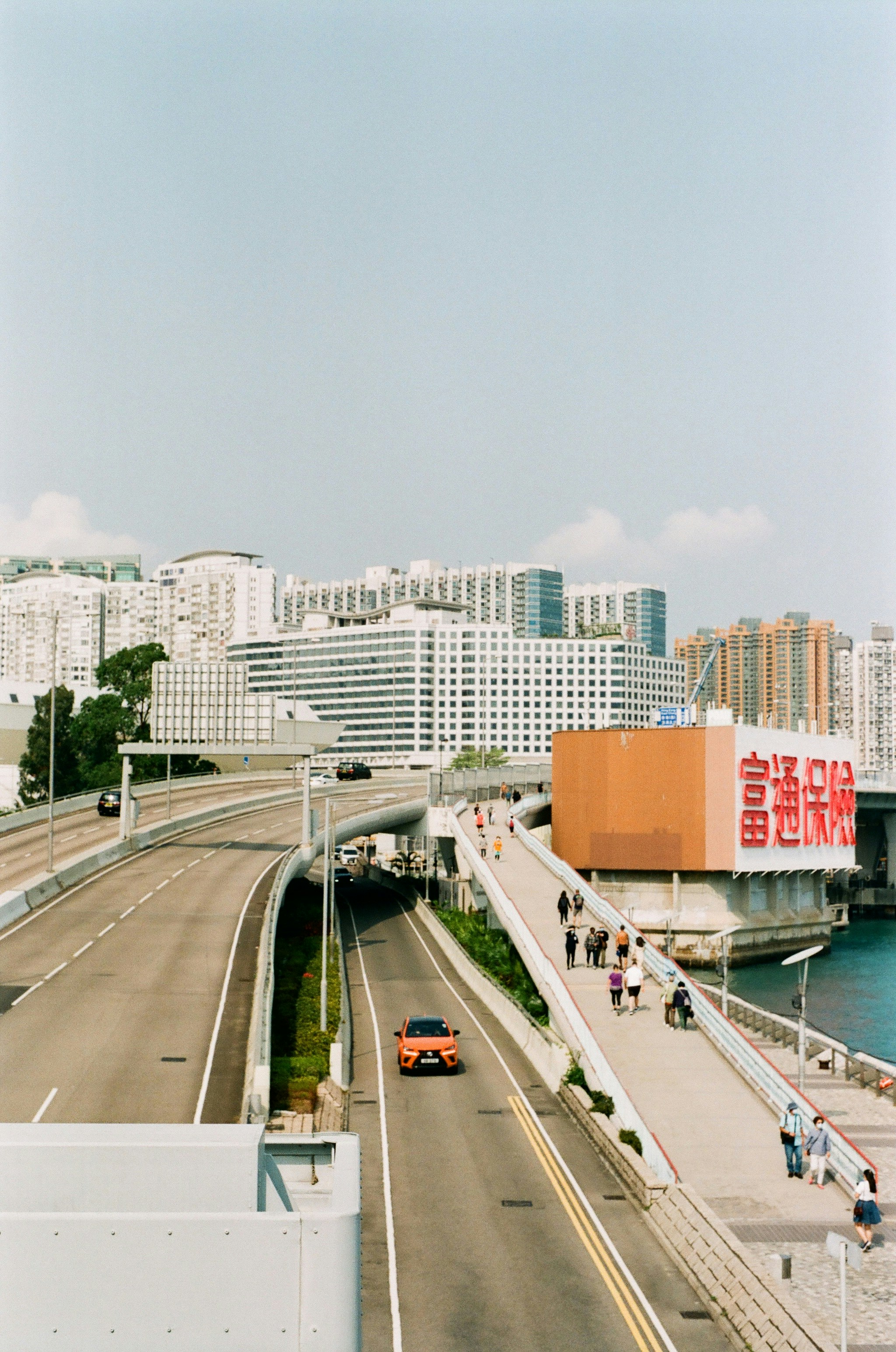A vibrant urban scene showcasing a bustling waterfront with pedestrians and vehicles, framed by modern architecture and a striking advertisement. 
