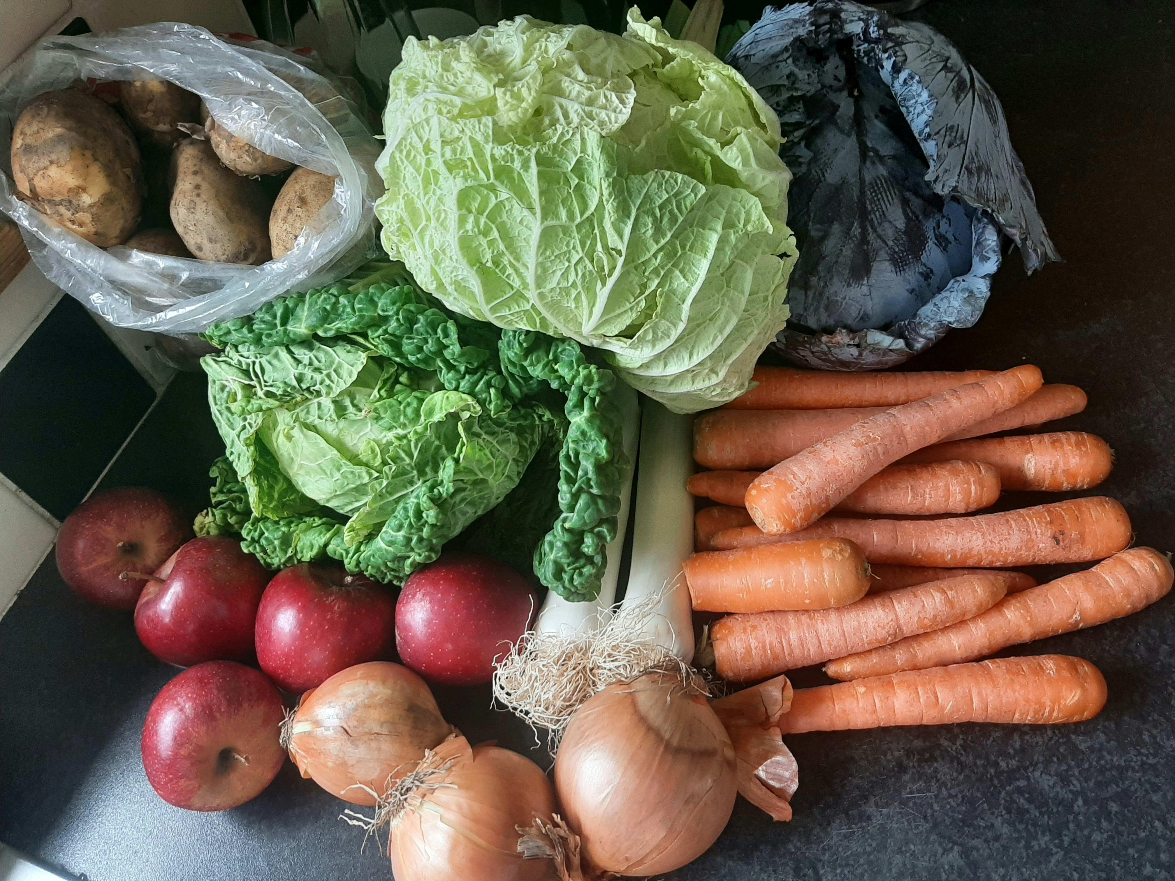 Photo of fresh vegetables arranged on a dark kitchen counter: carrots, lettuce, cabbage, onions, apples, and potatoes.