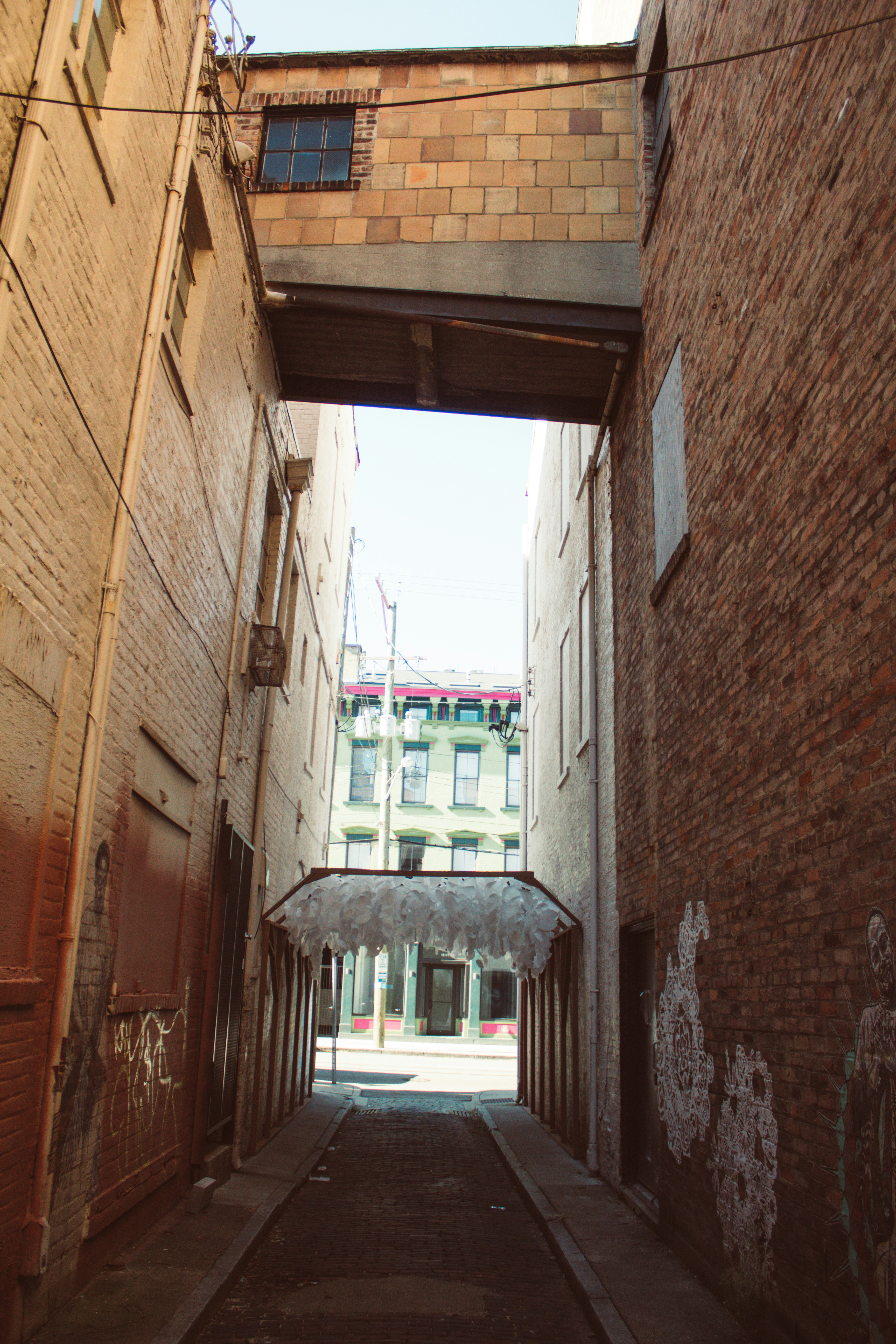 Narrow alleyway flanked by textured brick walls, featuring an overhead structure and glimpses of a vibrant building beyond.