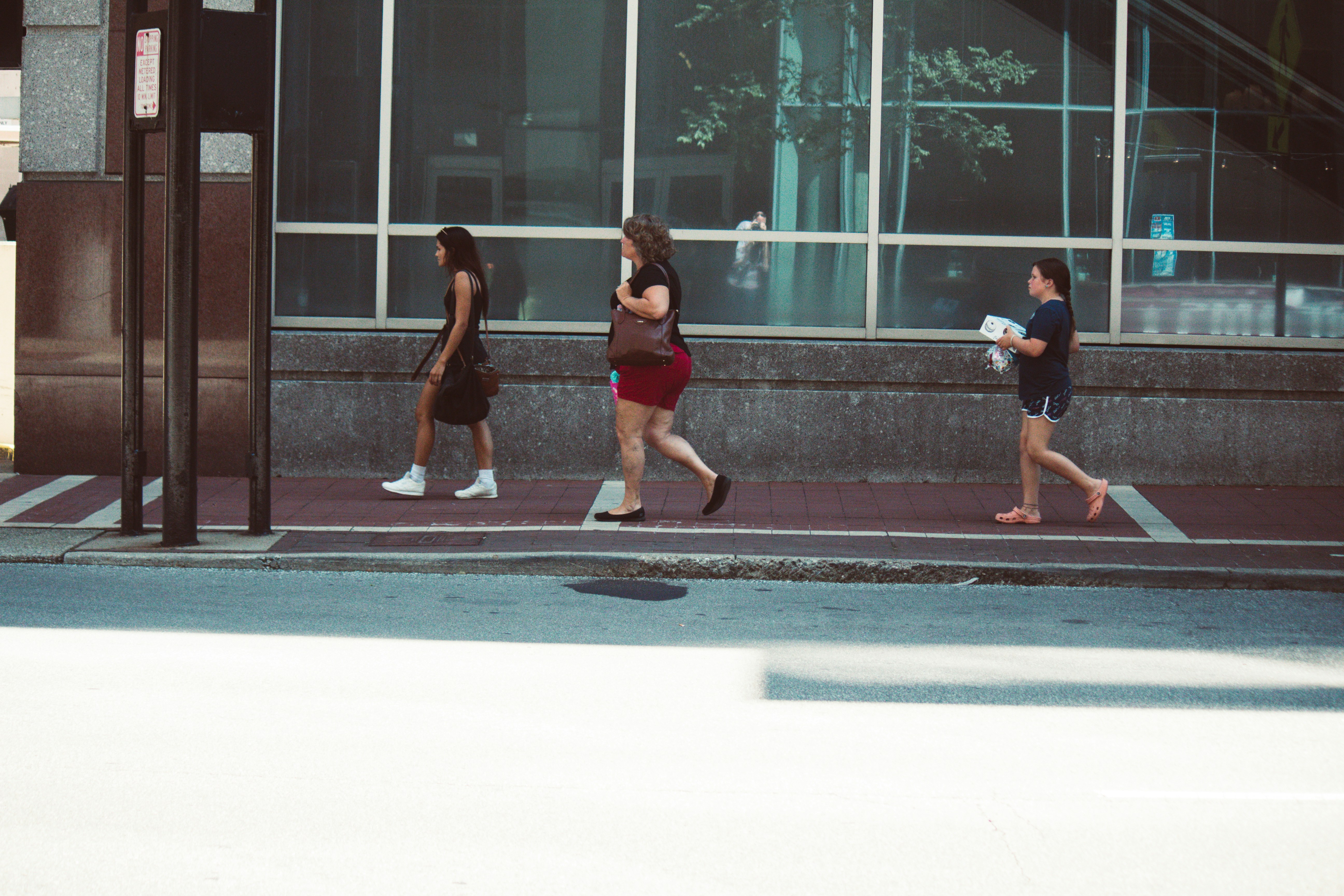 Three pedestrians walking along a city sidewalk, showcasing diverse styles and movements against a modern architectural backdrop.