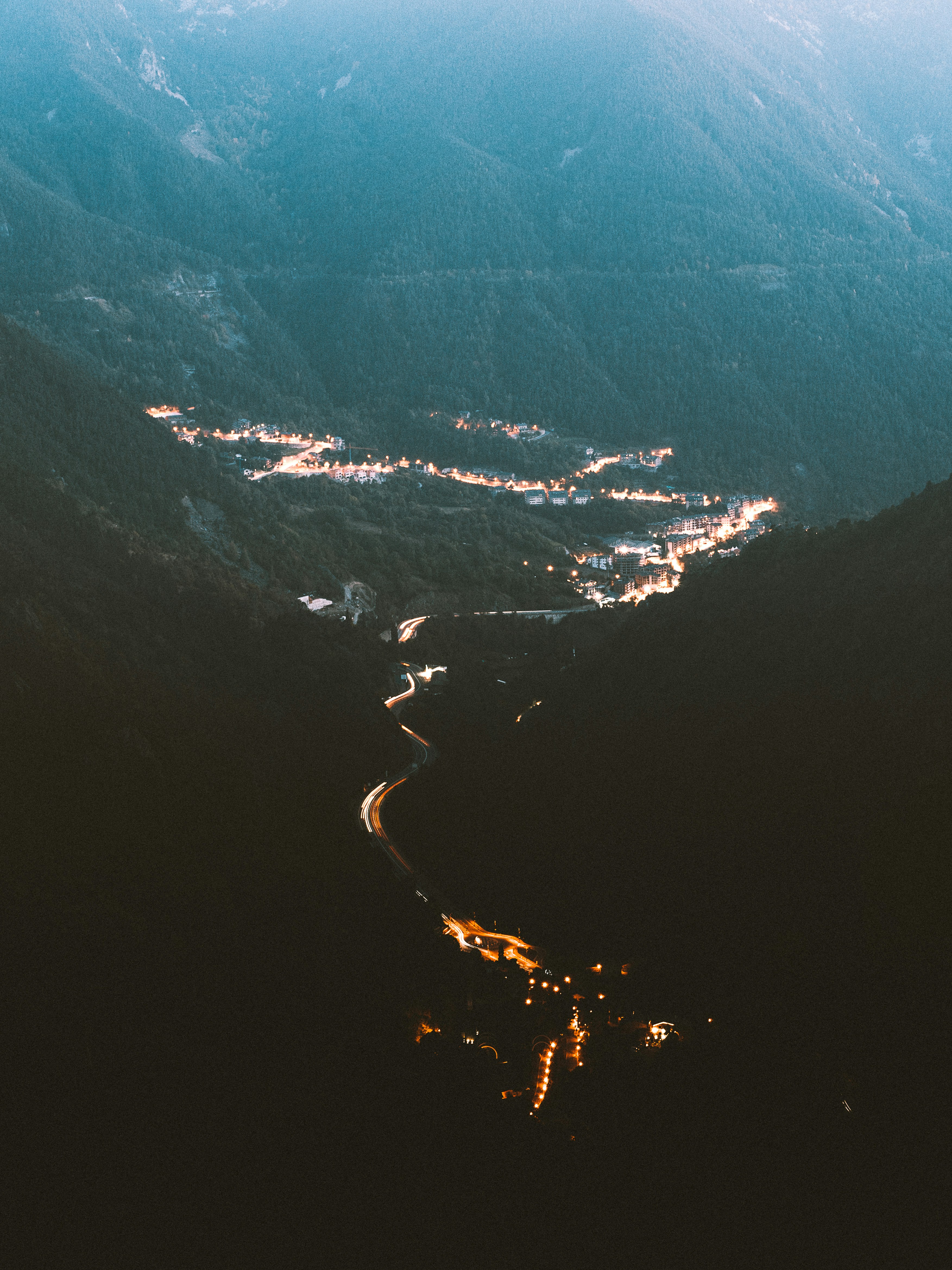 Aerial view of a mountain village illuminated at dusk, with winding roads and soft lights creating a tranquil atmosphere.