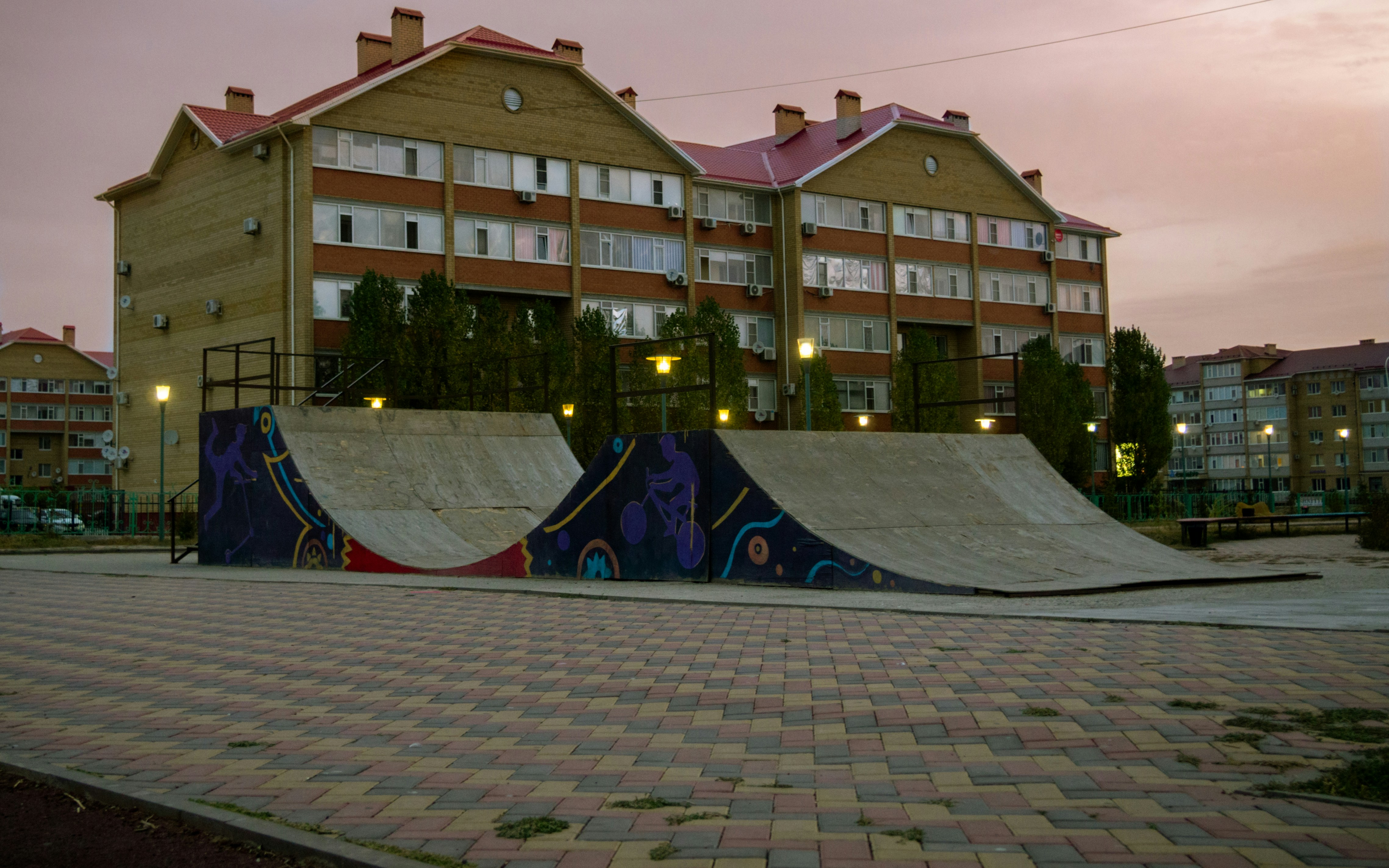 Photo of a graffiti-curved skate ramp in the foreground at dusk, with a row of apartment blocks lining the background. The wide tiled plaza foreground emphasizes the urban setting.