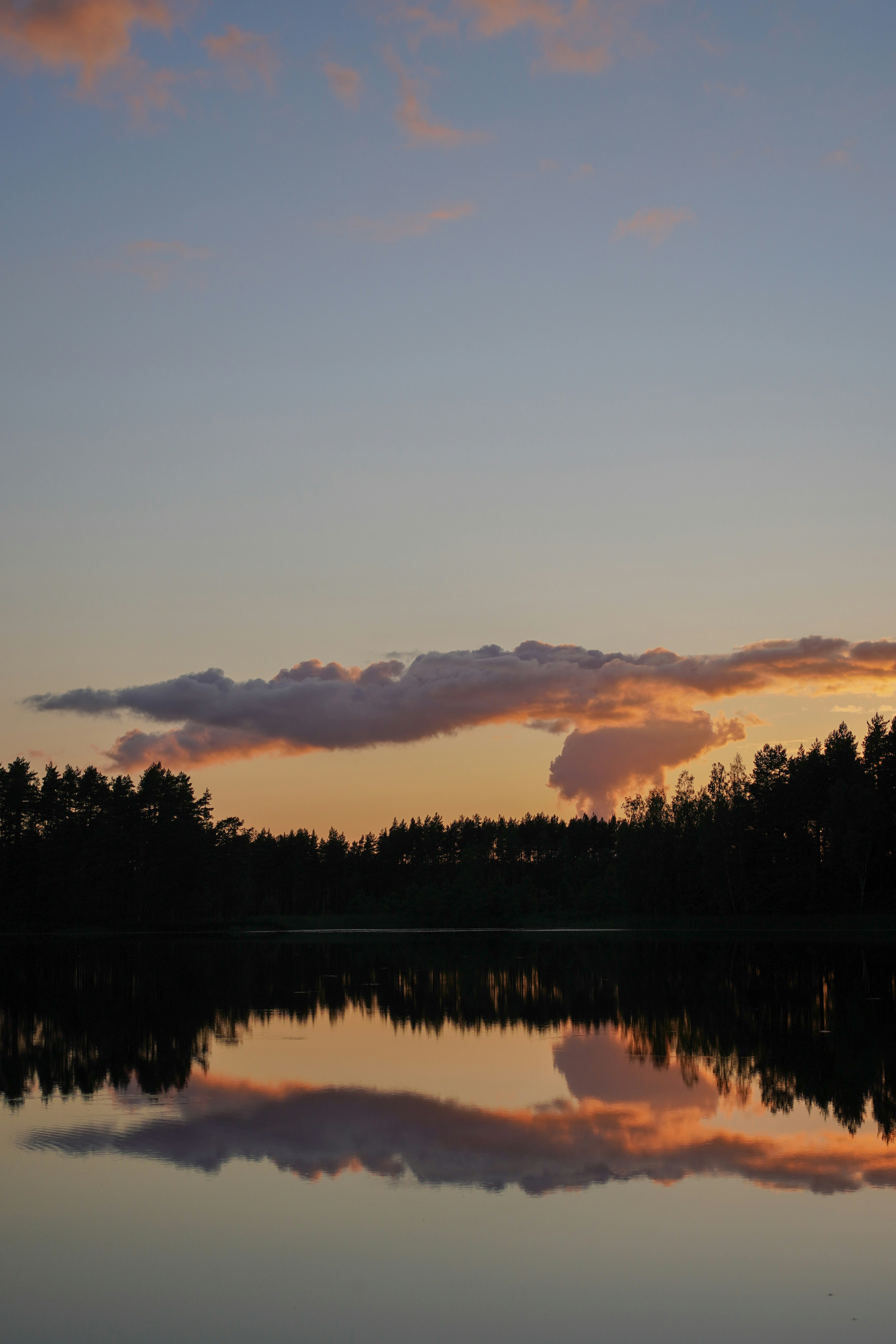 Serene lake reflecting a colorful sunset with soft clouds and silhouetted trees along the shore.
