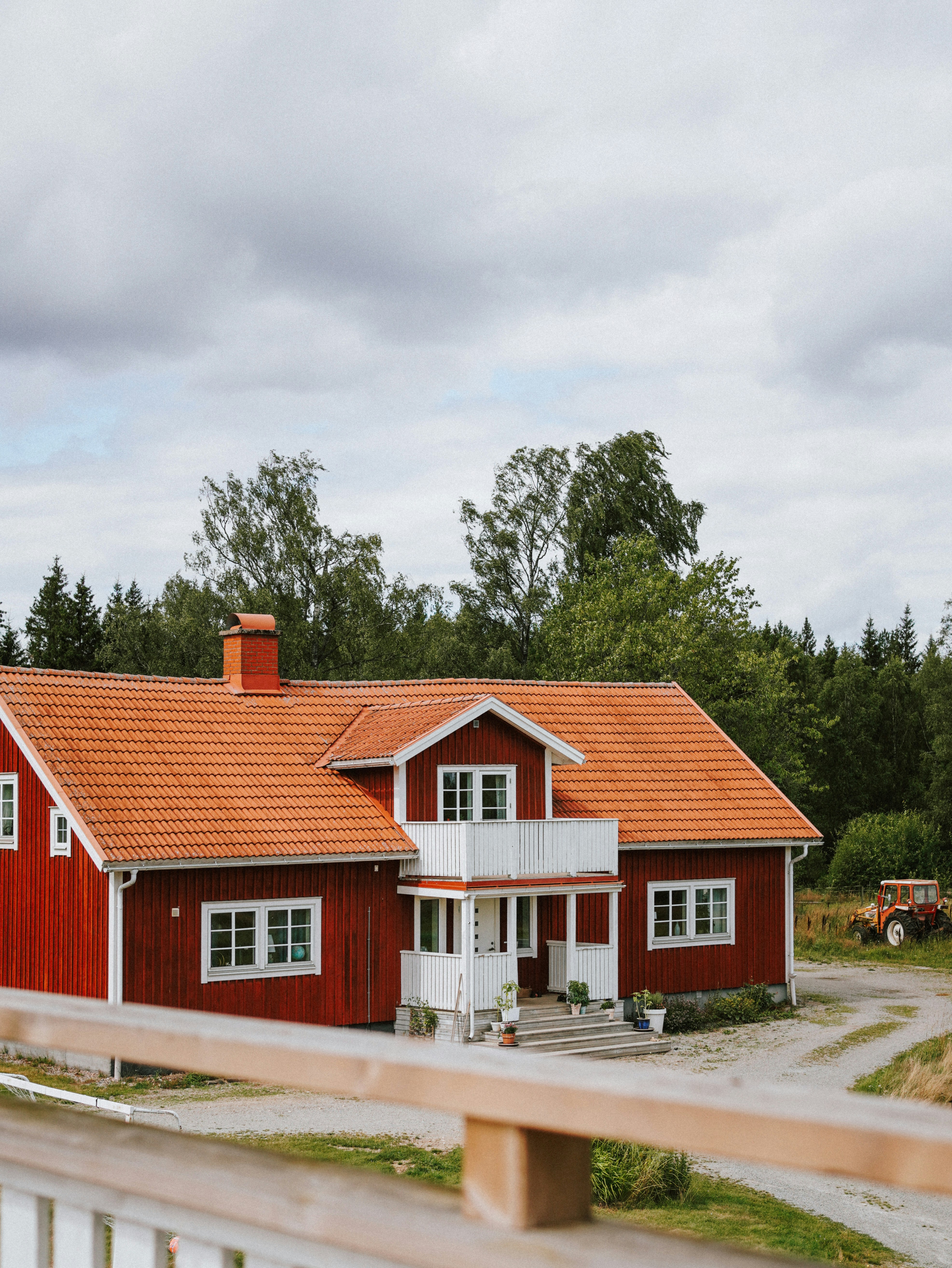 A vibrant red wooden house with a distinctive orange roof, surrounded by lush greenery and a gravel path, showcasing a tranquil countryside setting.