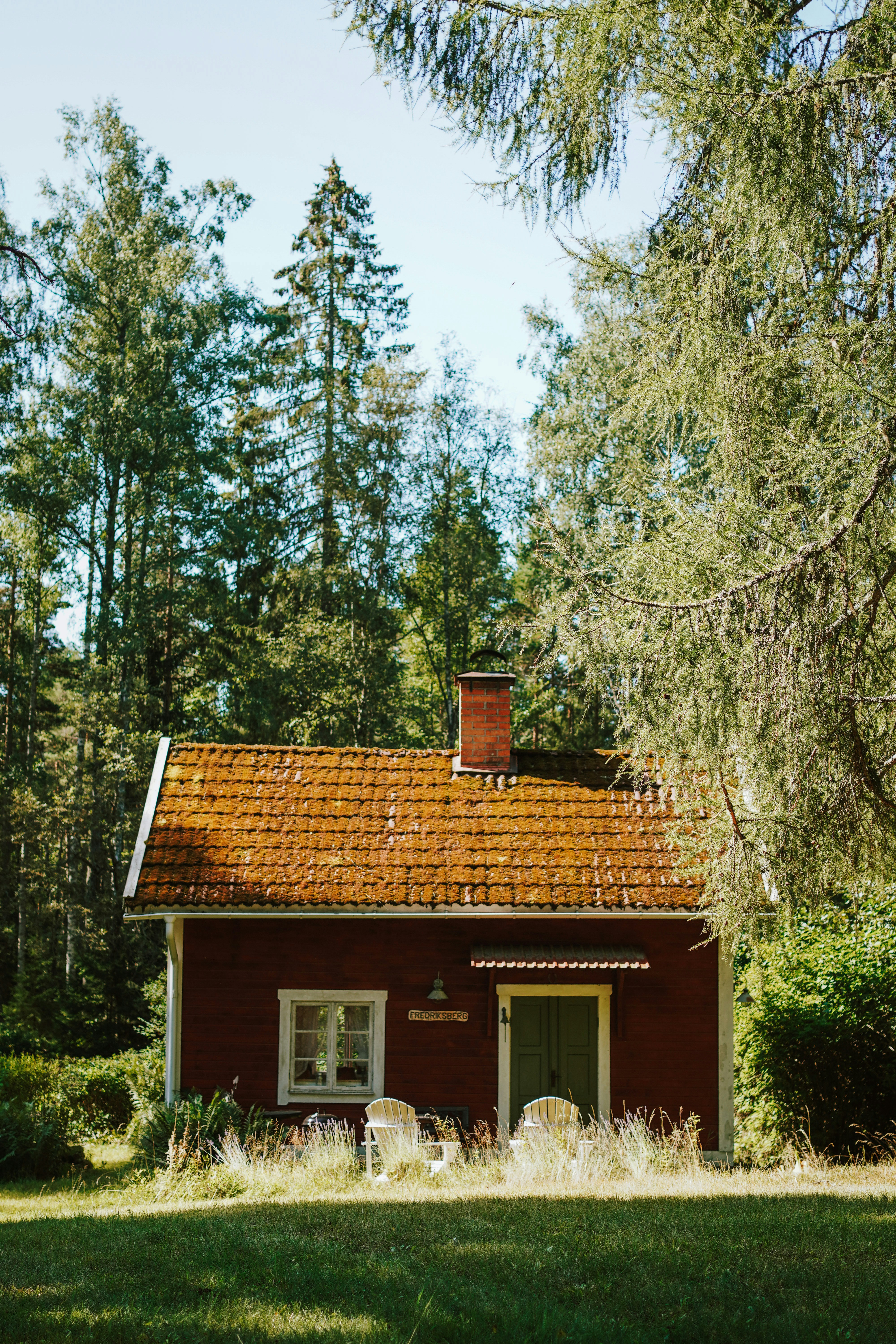 Charming red cabin nestled among lush green trees, featuring a moss-covered roof and inviting porch chairs. A serene escape into nature.