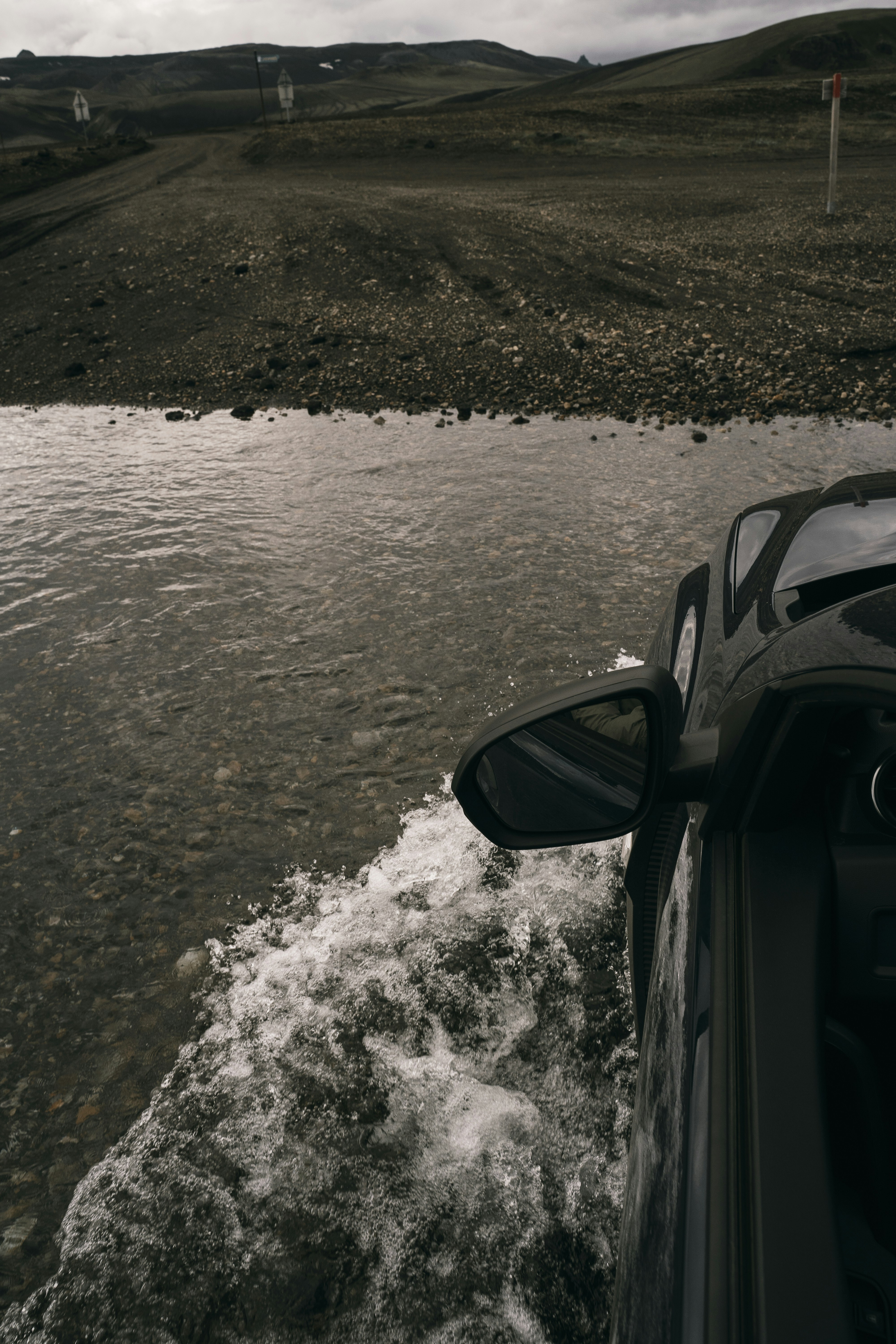 Riverside Skim: A Silver Car Slices a Pebble-Strewn Crossing Under Overcast Skies