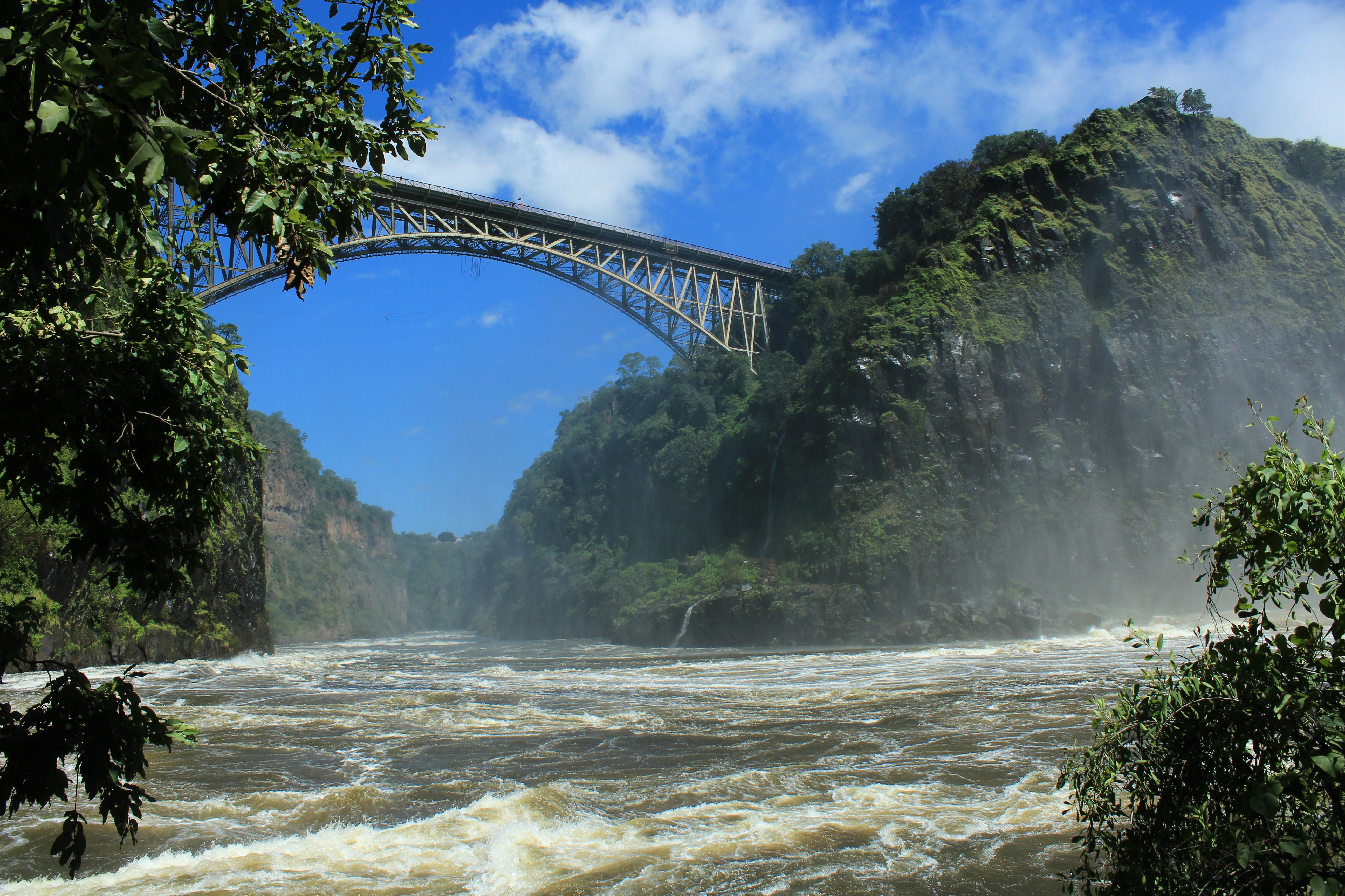 Steel arch bridge spanning over a turbulent river between lush, green cliffs under a bright blue sky.