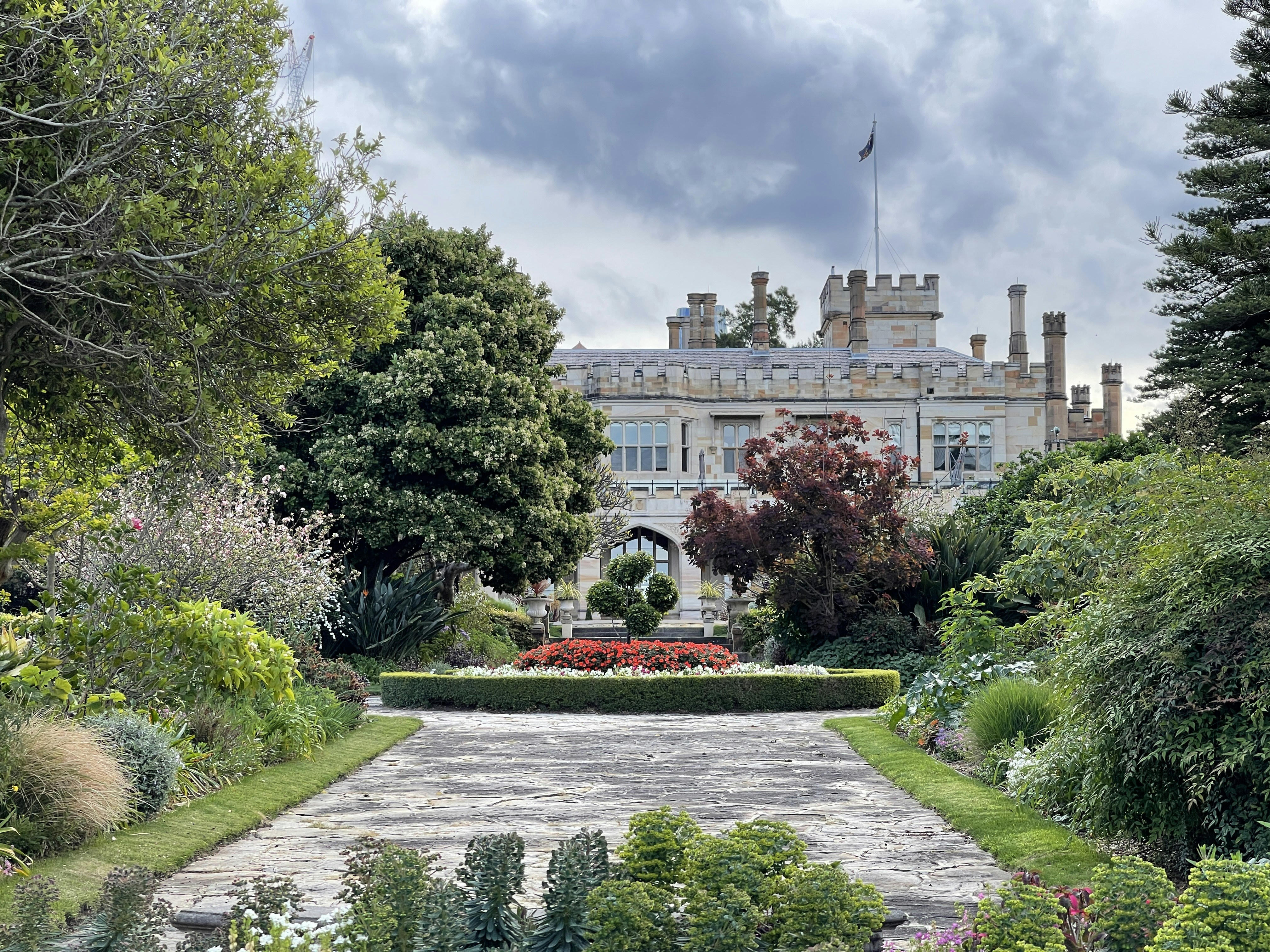 Lush garden path leads to a grand historic mansion, framed by vibrant flora and a dramatic sky.