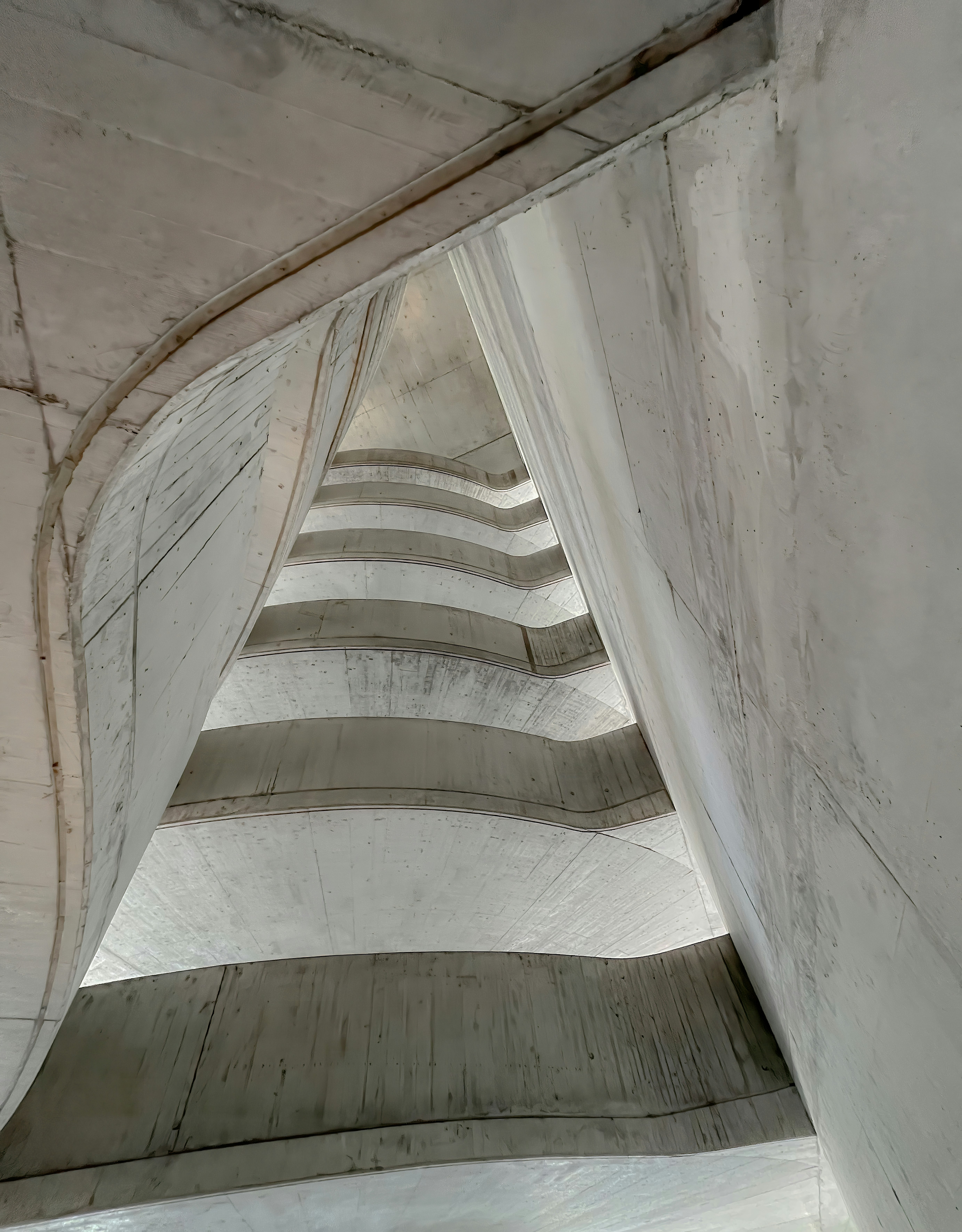 A sculptural concrete stairwell twists upward, creating a rhythm of curved ramps and pale gradients. The architectural interior is bathed in soft light that emphasizes the fluid planes.