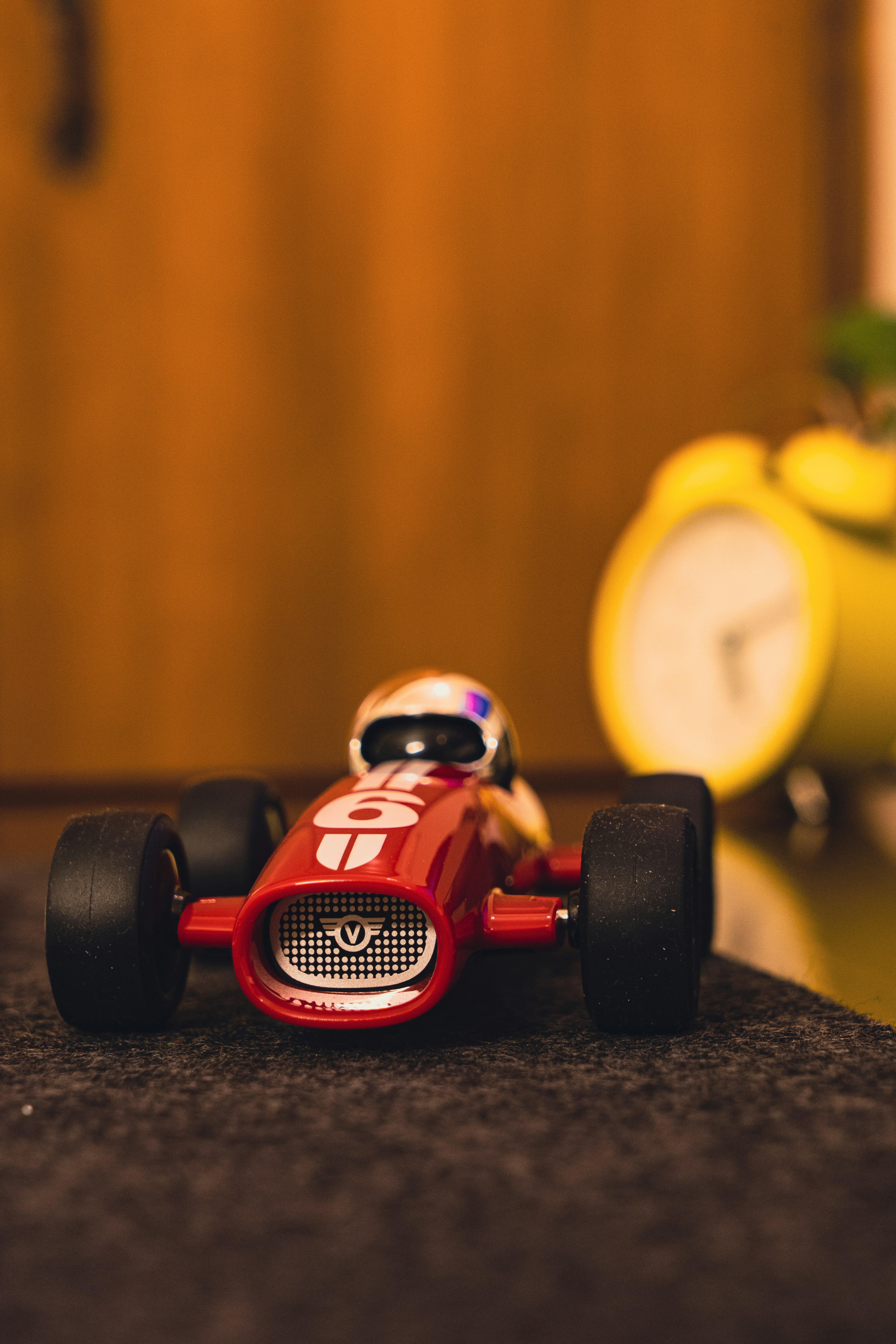 Red toy race car on a soft dark surface with a yellow alarm clock in the blurred background