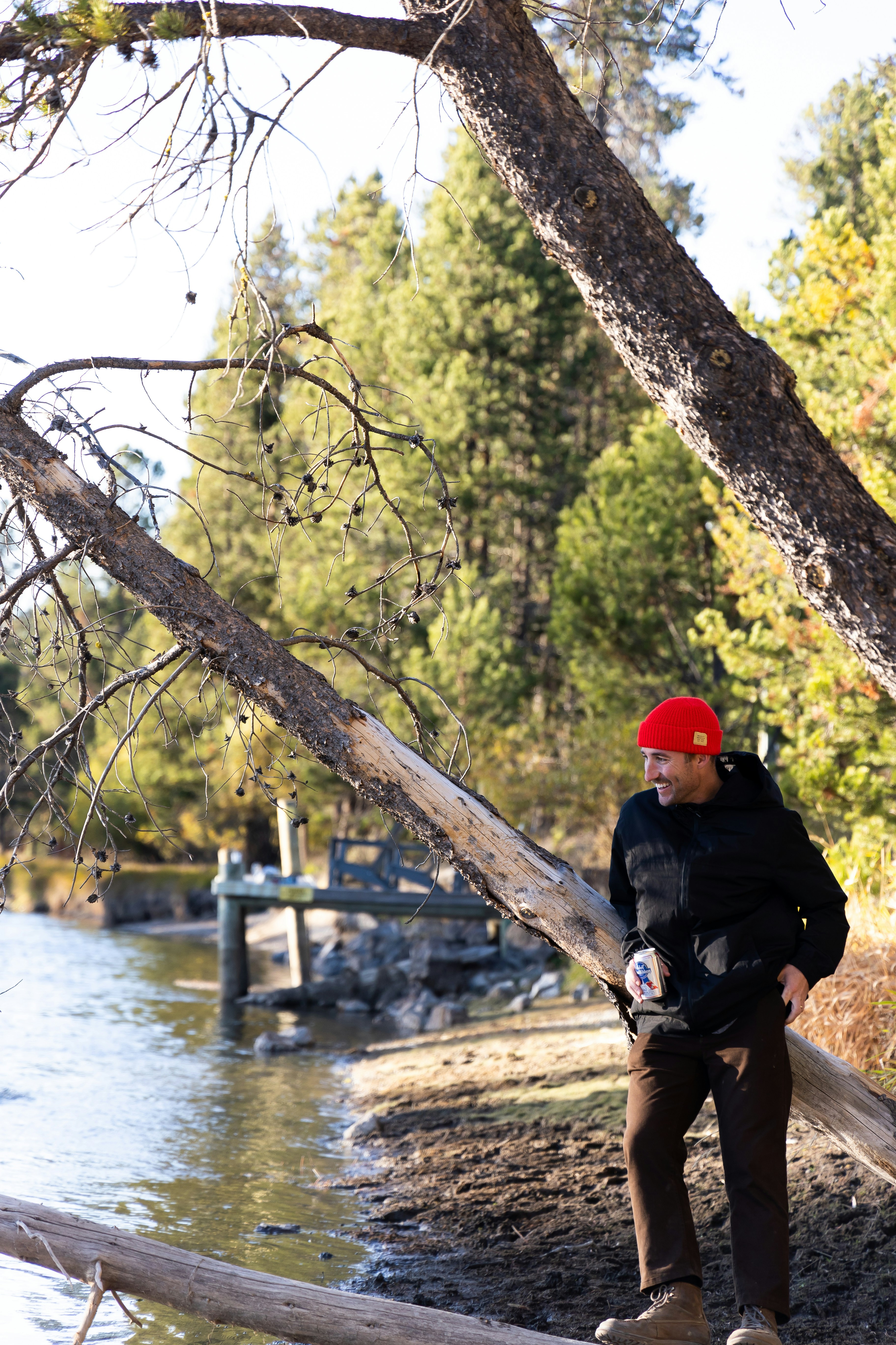 Man in a red beanie enjoying a drink while sitting by a serene lakeside, surrounded by lush greenery and fallen branches.