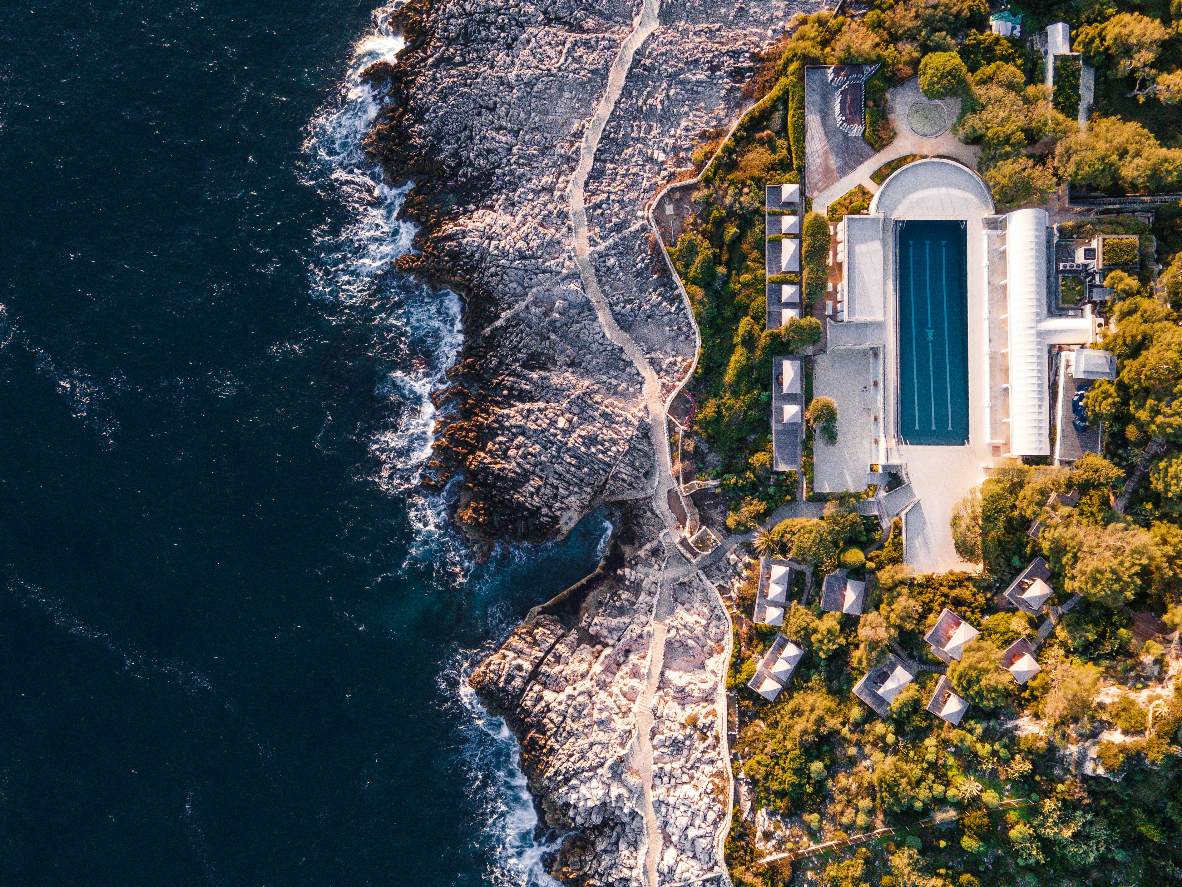 Aerial view of a coastal retreat featuring a swimming pool surrounded by lush greenery and rocky shoreline. The image captures the harmonious blend of nature and architecture.