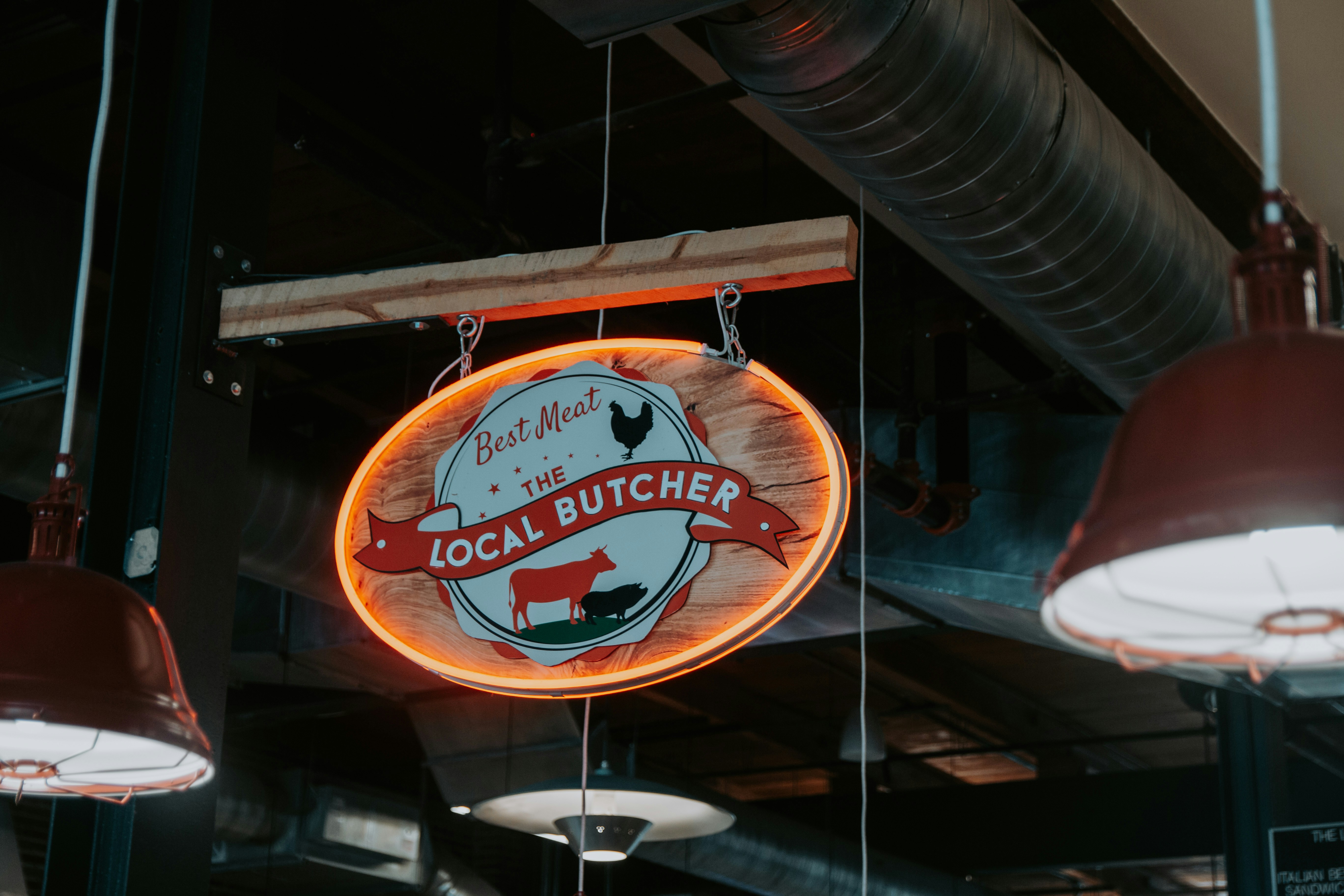 Neon-lit sign for 'The Local Butcher' featuring a cow and chicken, suspended in a rustic market setting.