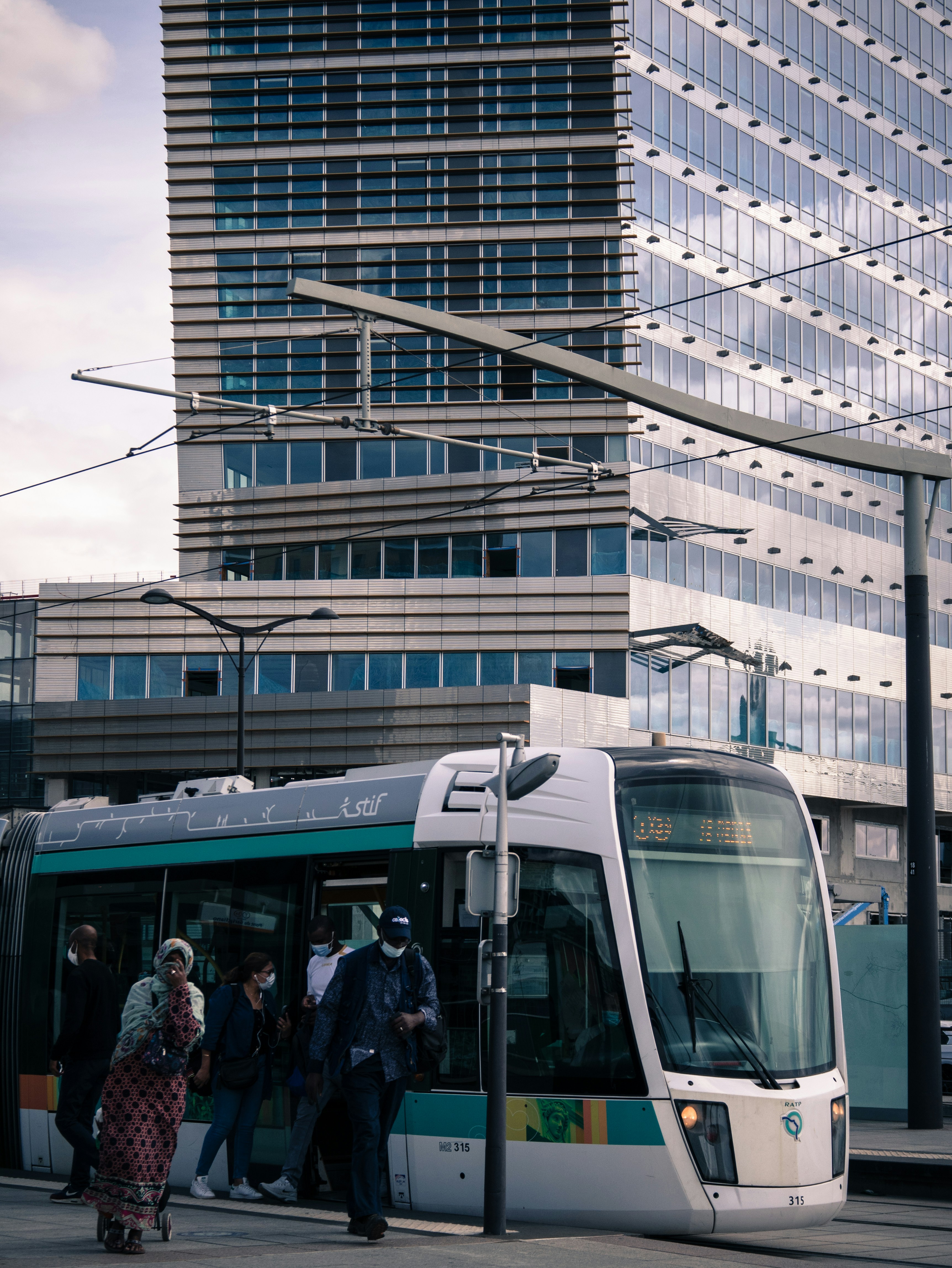 Tram station bustling with commuters as a modern tram arrives, framed by contemporary architecture. The scene captures the essence of urban mobility.