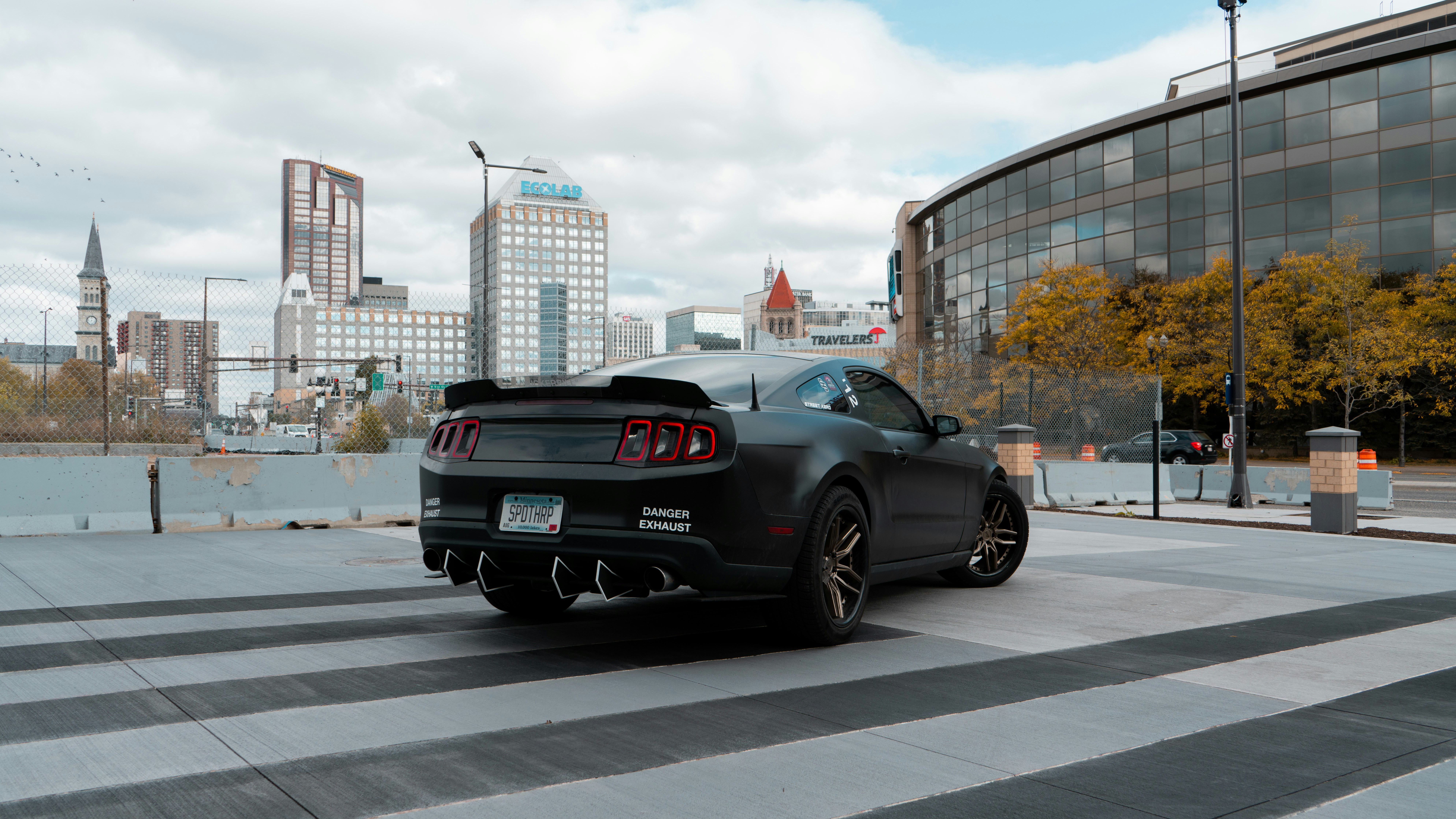 Matte black Mustang parked on a city street, showcasing its aggressive rear design against an urban skyline. The backdrop features modern buildings and autumn foliage.
