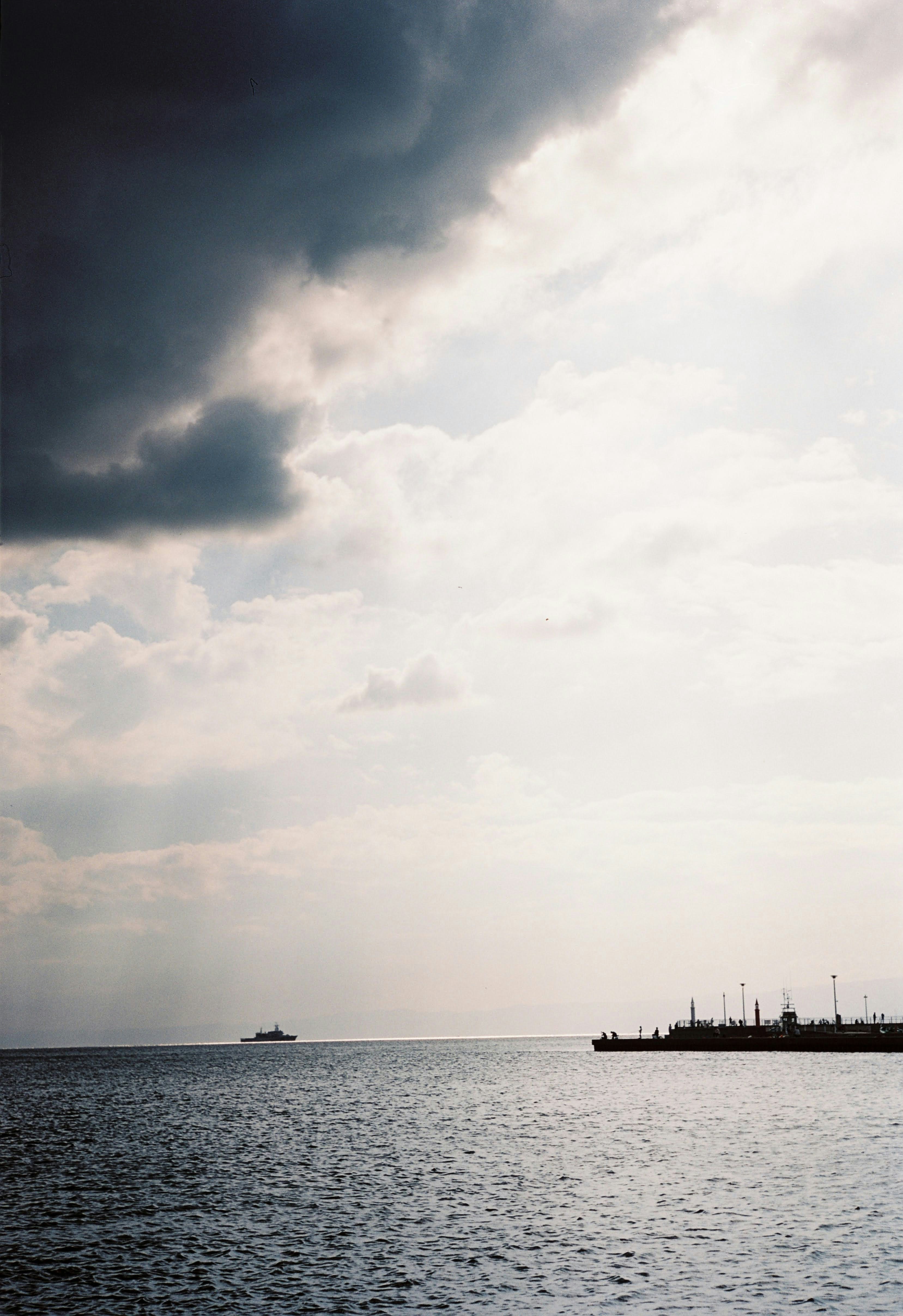 A tranquil harbor scene featuring a distant ship and a pier against a backdrop of dramatic clouds. The calm waters reflect the shifting light of the overcast sky.