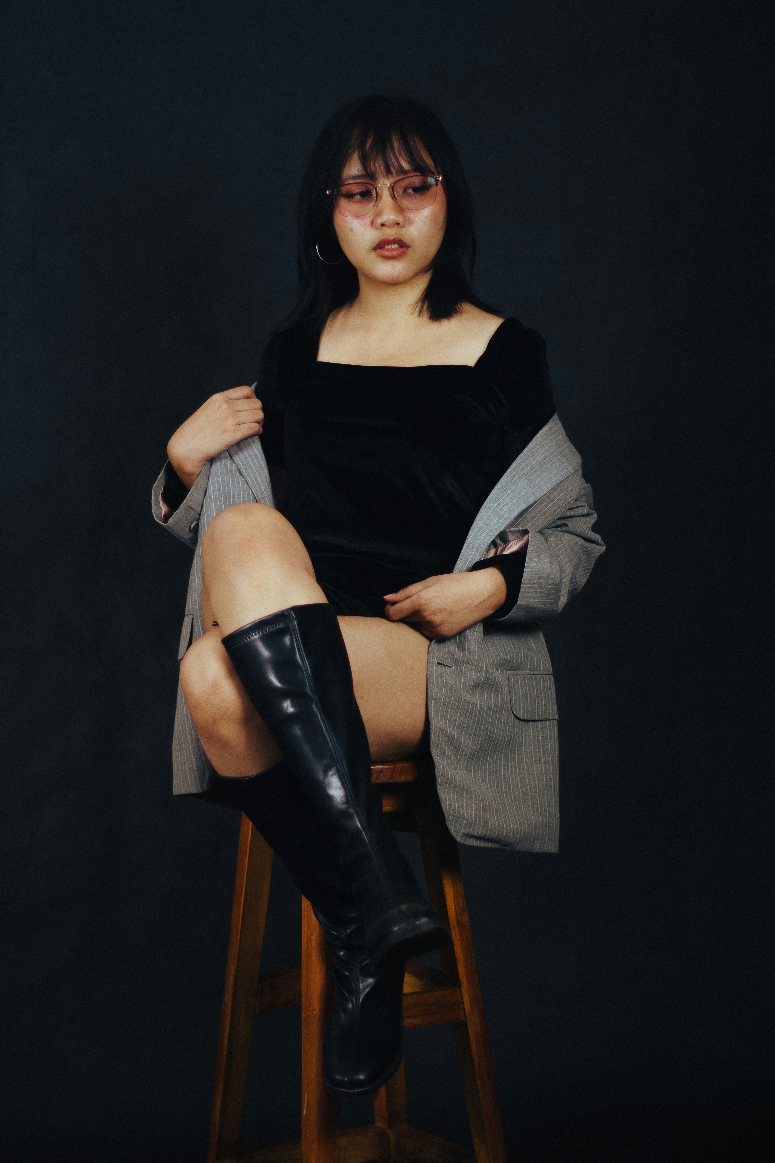 A fashion-forward individual poses confidently on a stool, dressed in a black velvet outfit and oversized blazer, set against a dark backdrop.