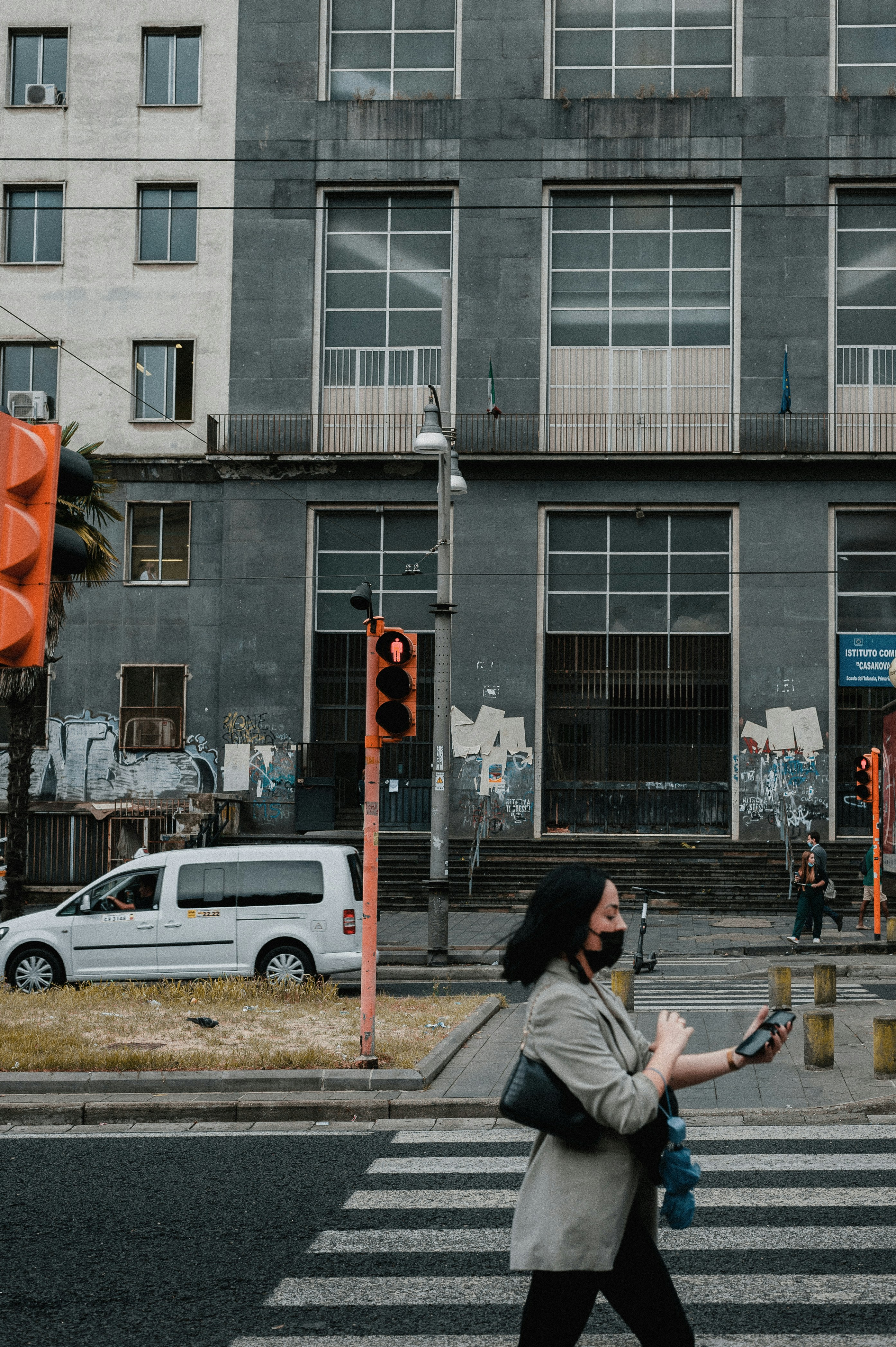 A woman walks across a crosswalk in front of a weathered building, with traffic lights and a van in the background.