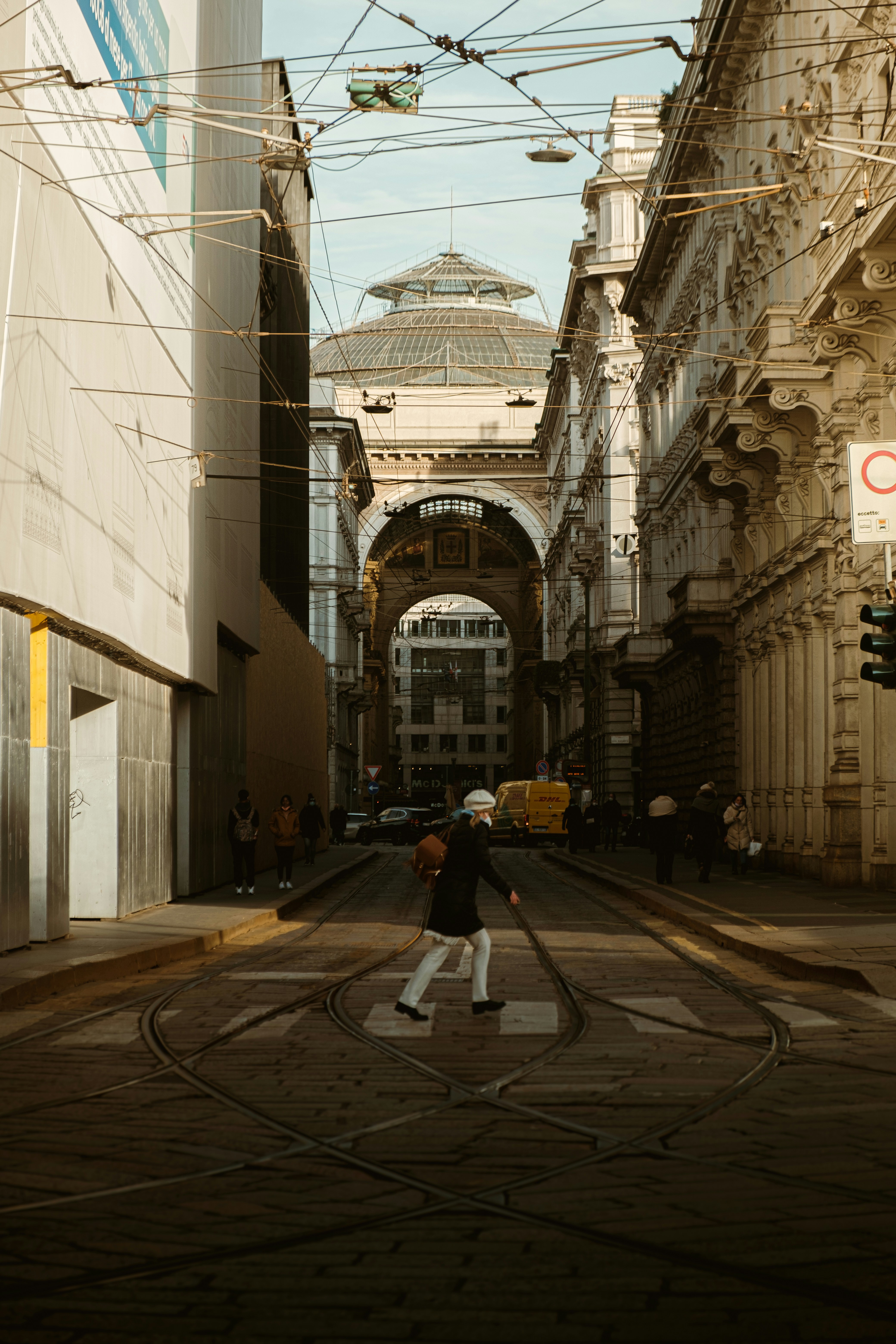 A pedestrian crosses tram tracks in a narrow city street, framed by grand architectural details and soft morning light.