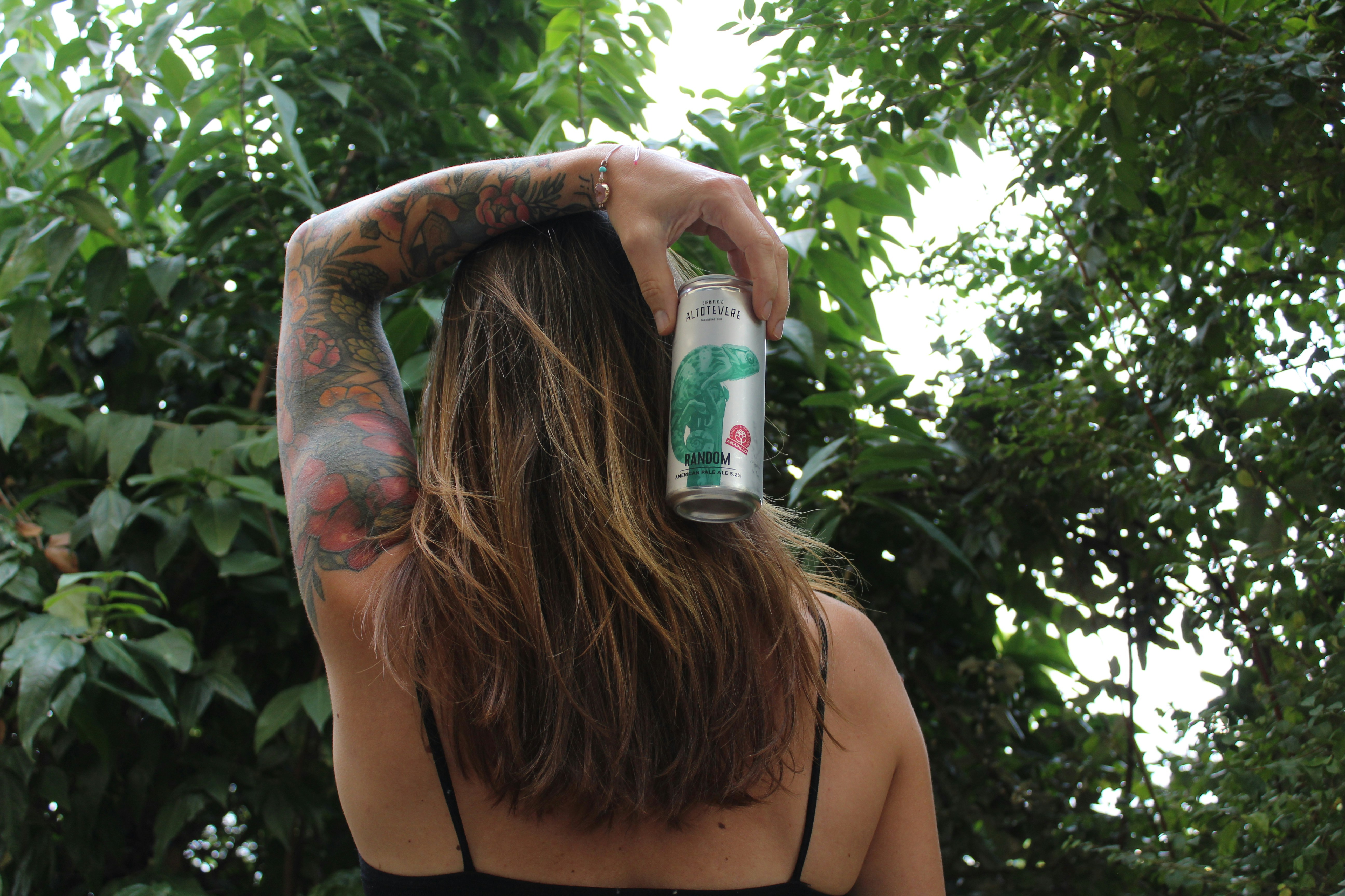 Woman with colorful tattoos holds a beverage can above her head against a lush green backdrop.