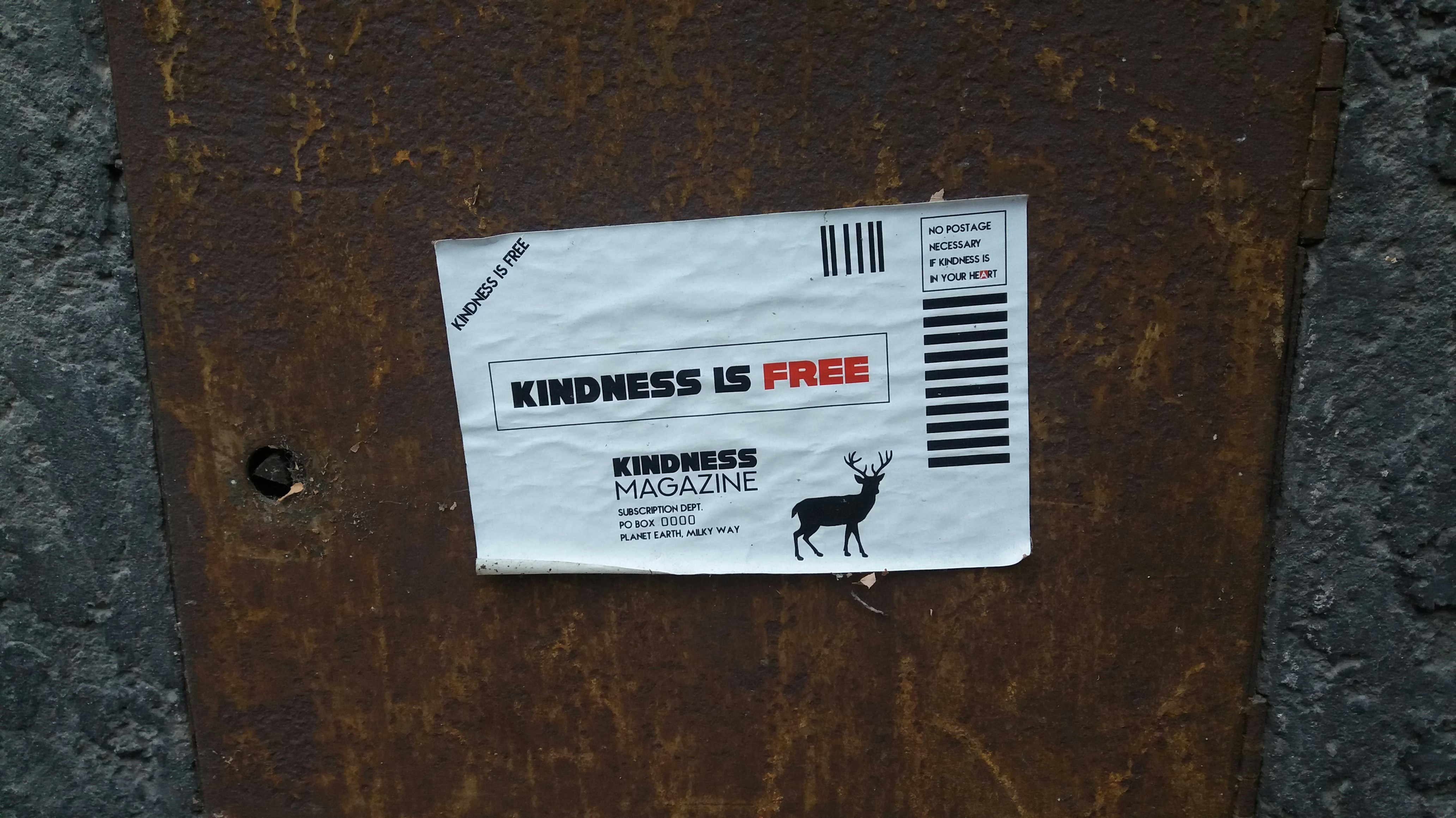 White sticker reading 'KINDNESS IS FREE' from Kindness Magazine is affixed to a weathered rusted metal surface, featuring a small deer logo.