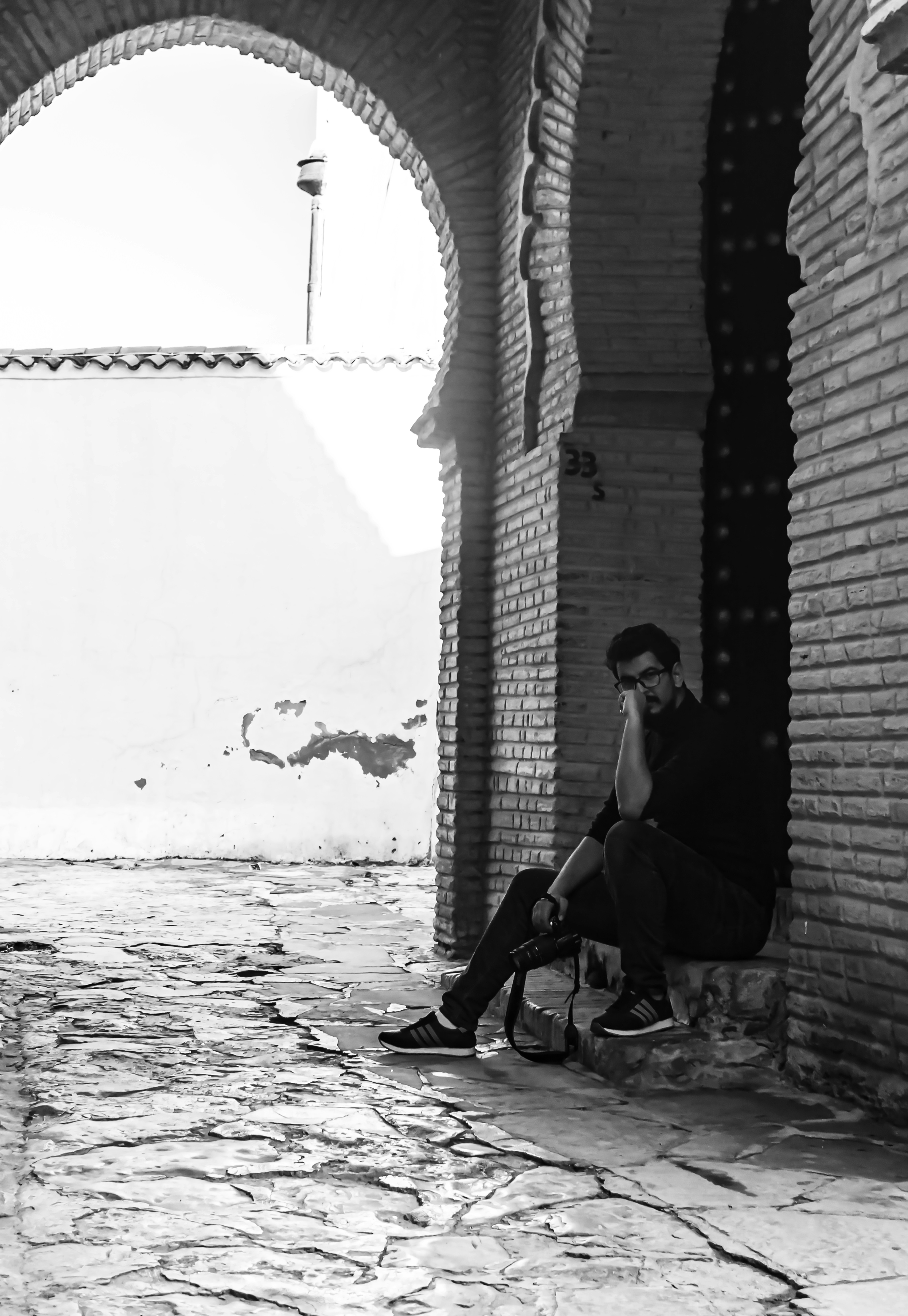A lone figure seated in a sunlit corridor, deep in thought, surrounded by textured brick walls and archways.