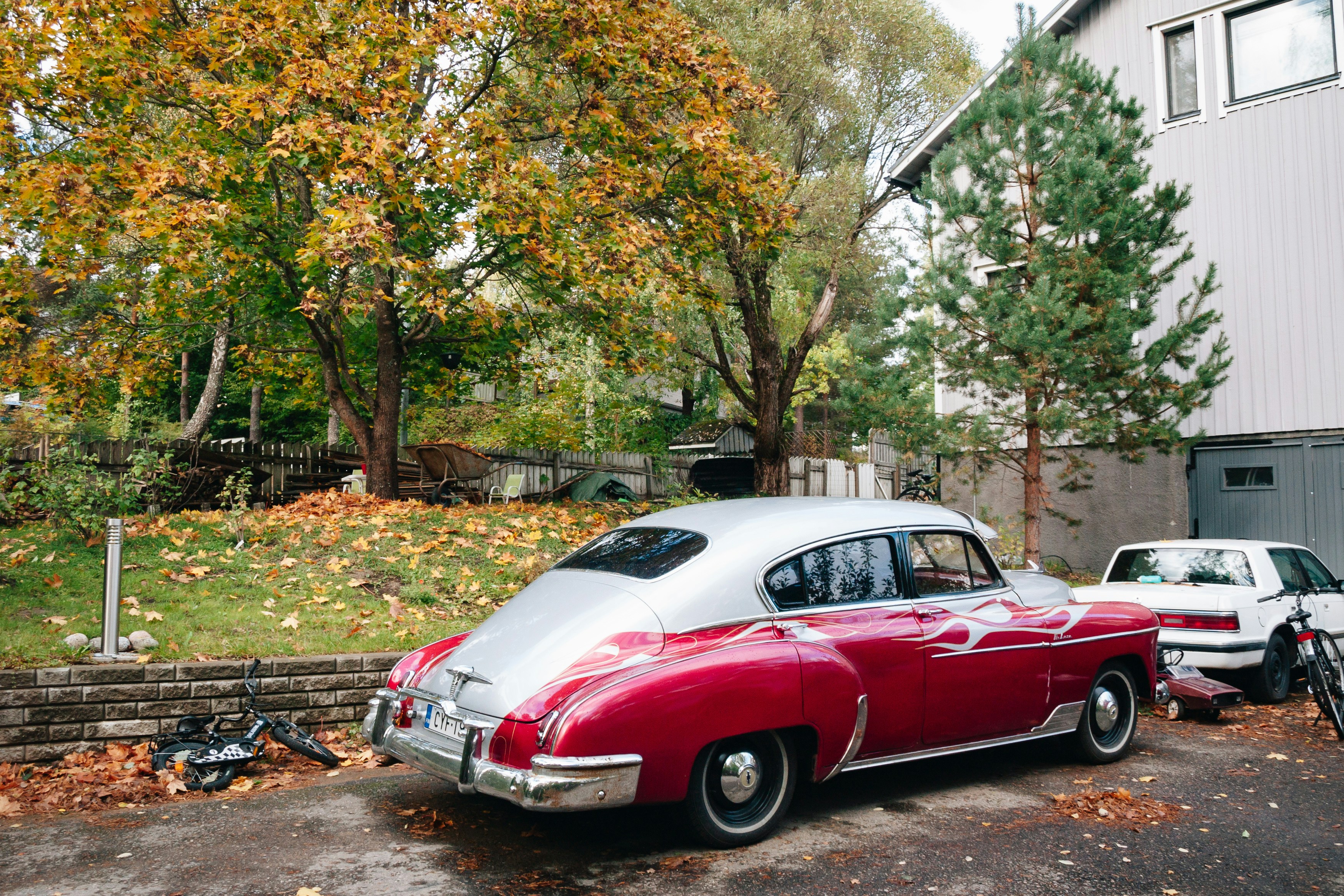 A glossy red vintage car parked on a wet street with autumn leaves and a gray building in the background.