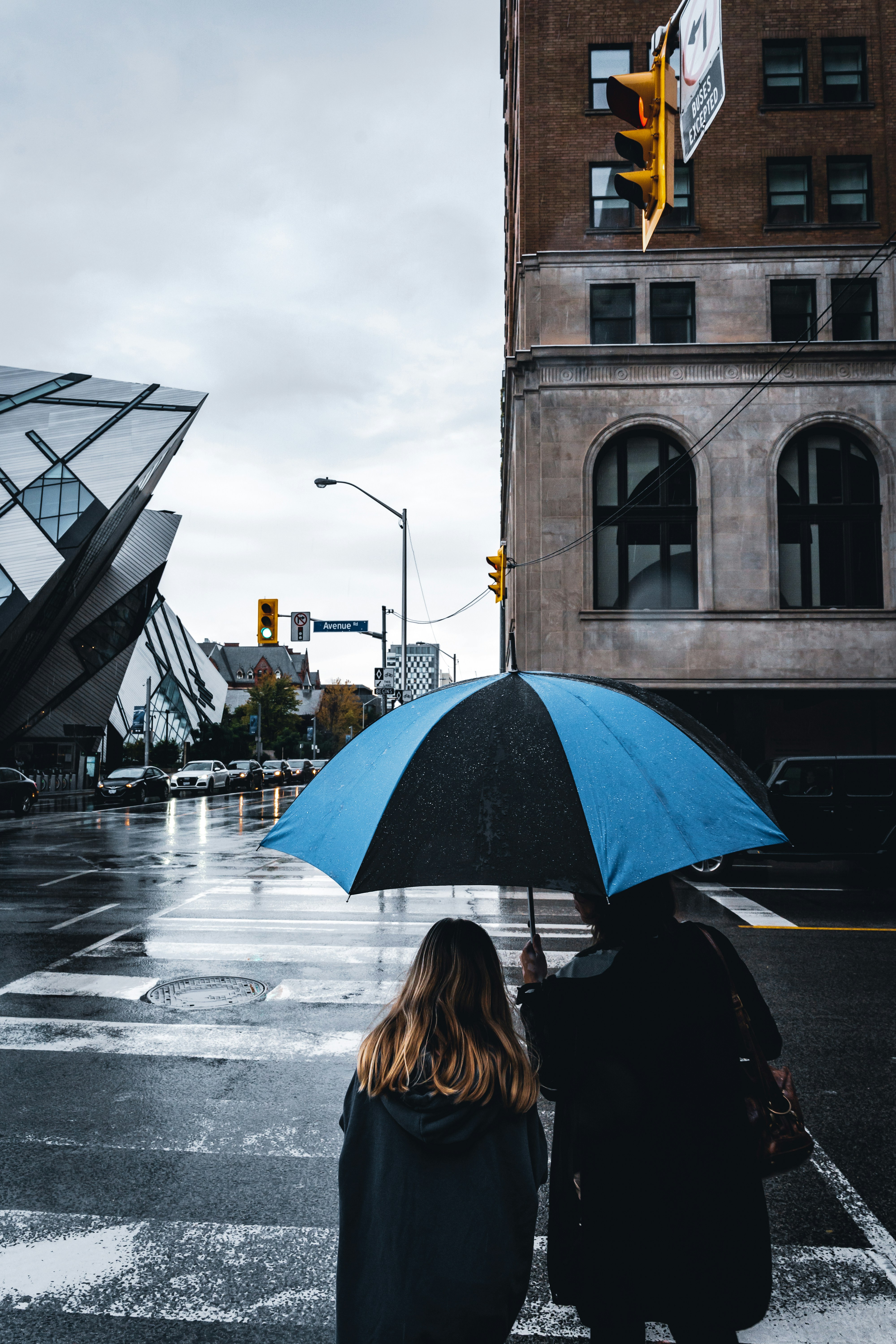 Two figures share a blue umbrella while crossing a city street, surrounded by modern architecture and a moody sky. Rain droplets glisten on the pavement.