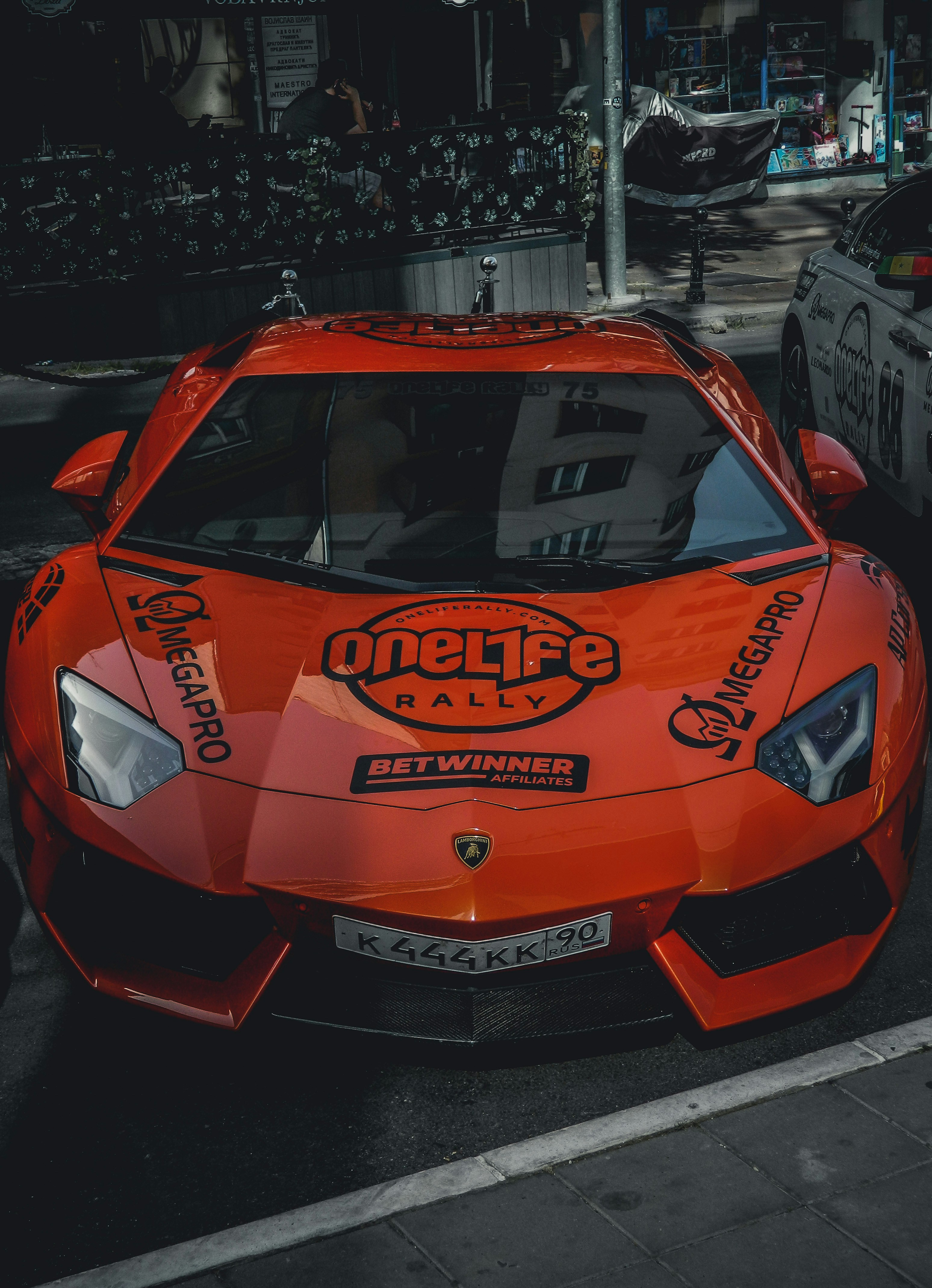 Photograph of a bright orange Lamborghini Huracán with One Life Rally branding on the hood, parked on a city street.