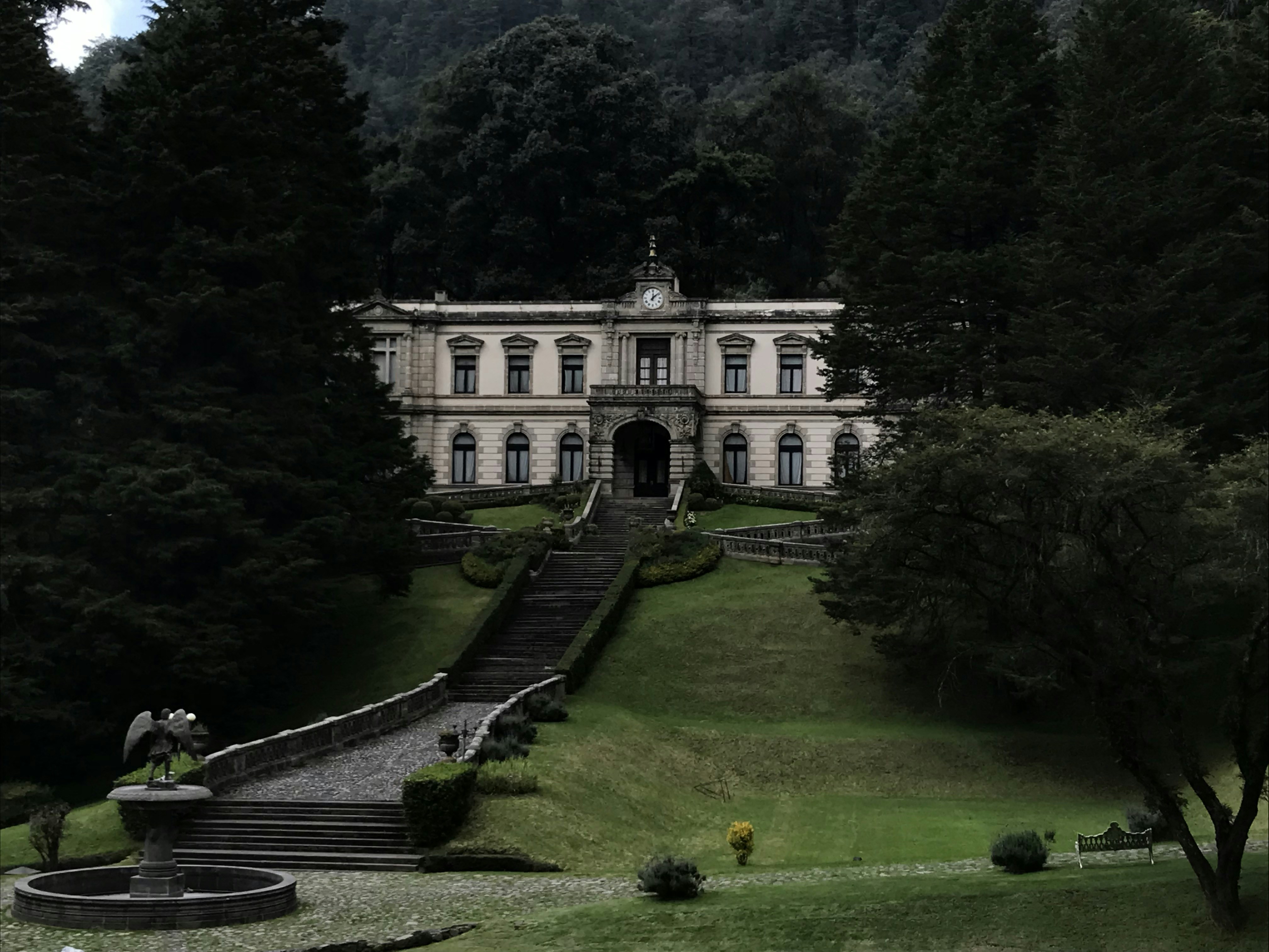 Grand mansion nestled among lush greenery, featuring a central staircase leading to an ornate entrance. The serene landscape enhances the architectural beauty.