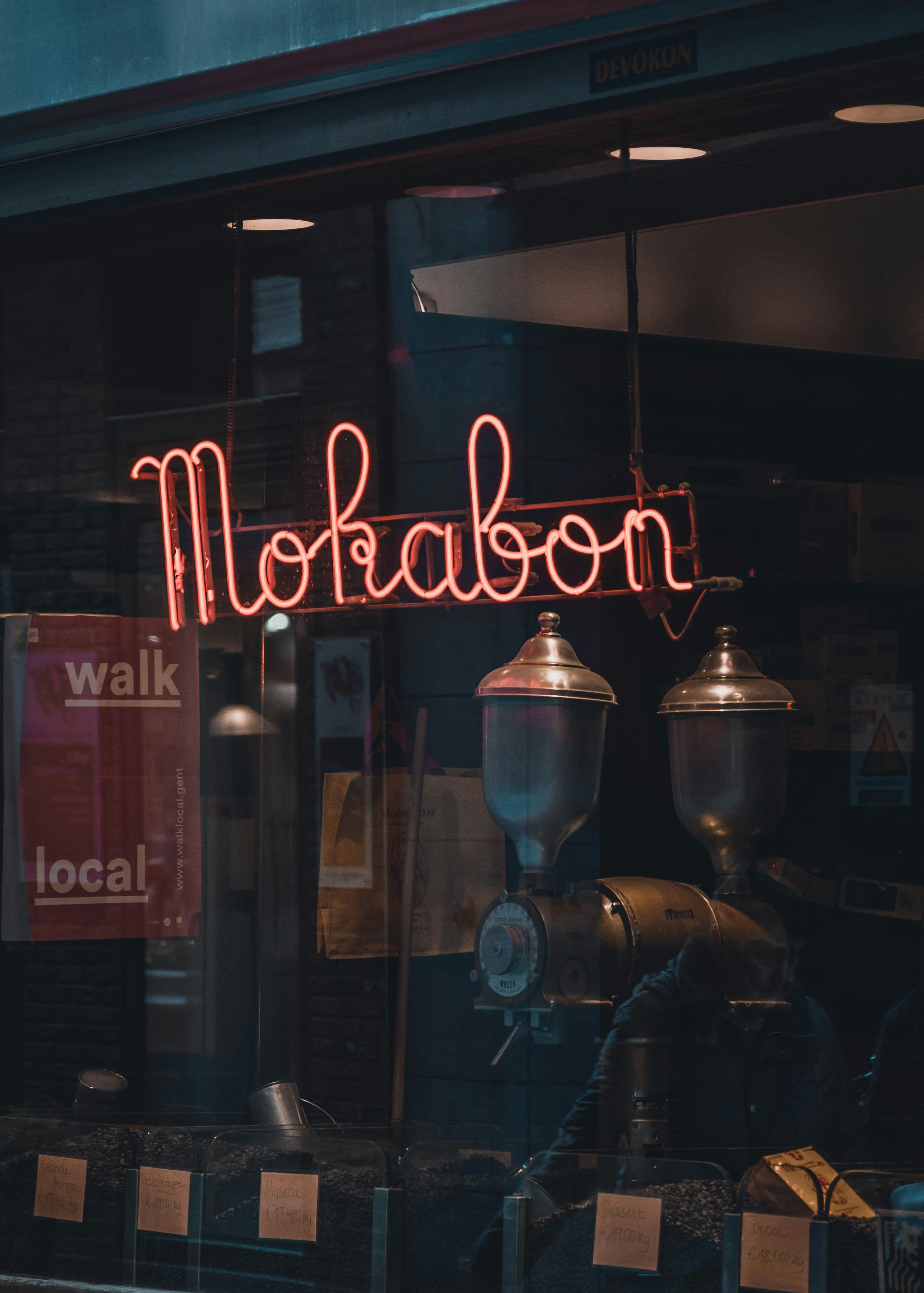 Neon sign reading 'Mokabon' glows warmly in a café window, highlighting brewing equipment and inviting ambiance.