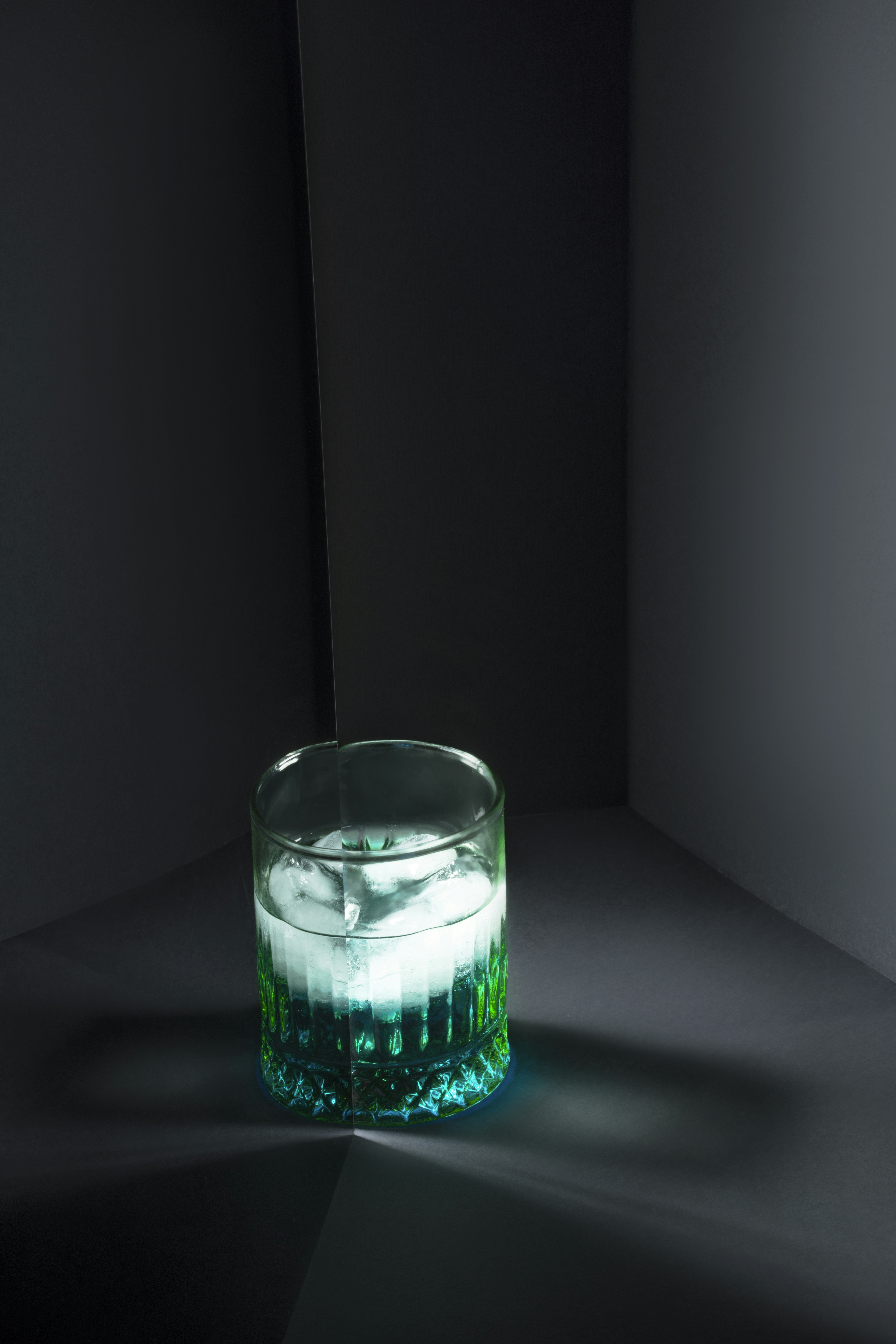 A crystal glass filled with ice and a green liquid rests in a dark corner, illuminated softly to highlight its contours and reflections.