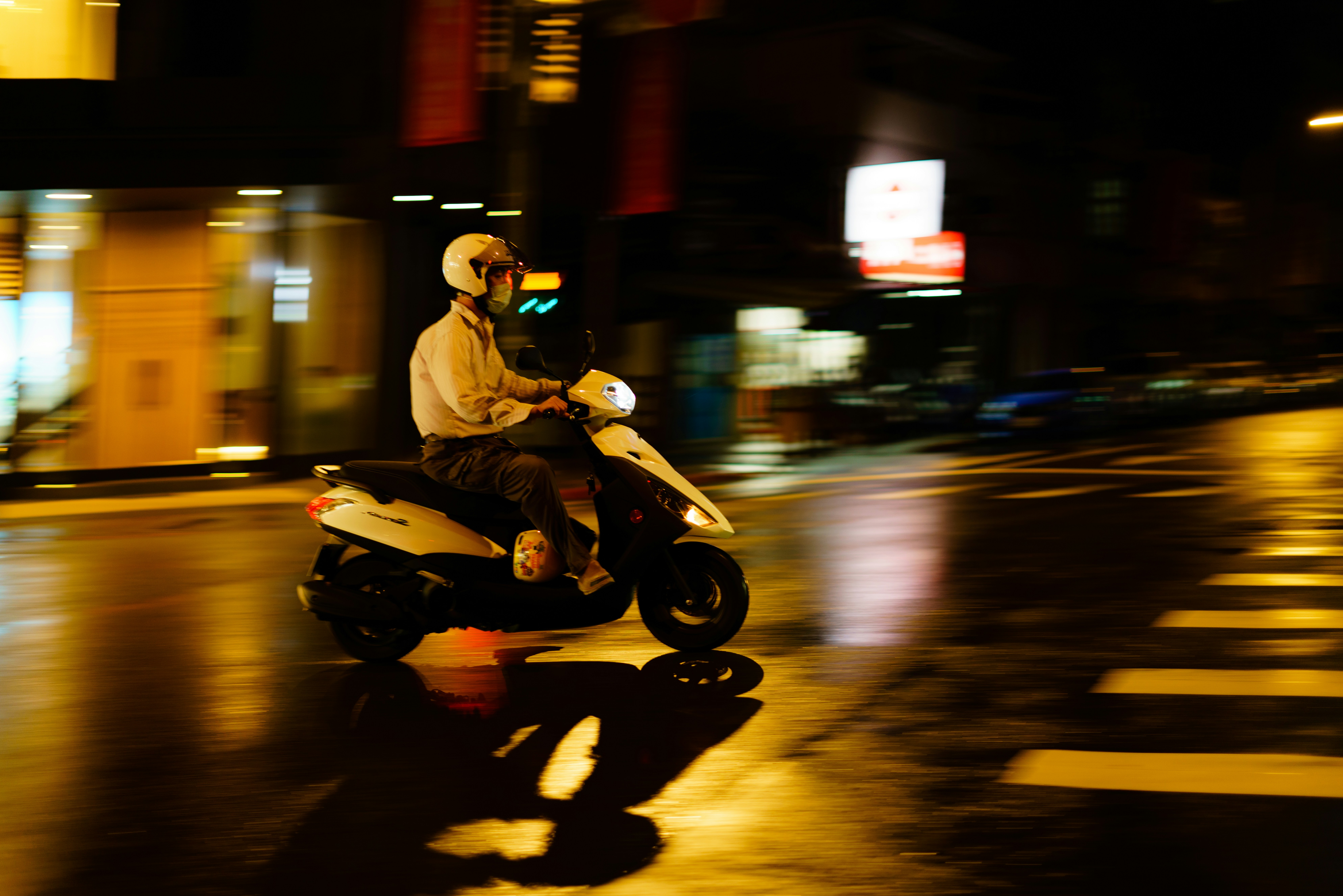 A scooter rider navigates a slick city street at night, illuminated by vibrant reflections and urban lights.