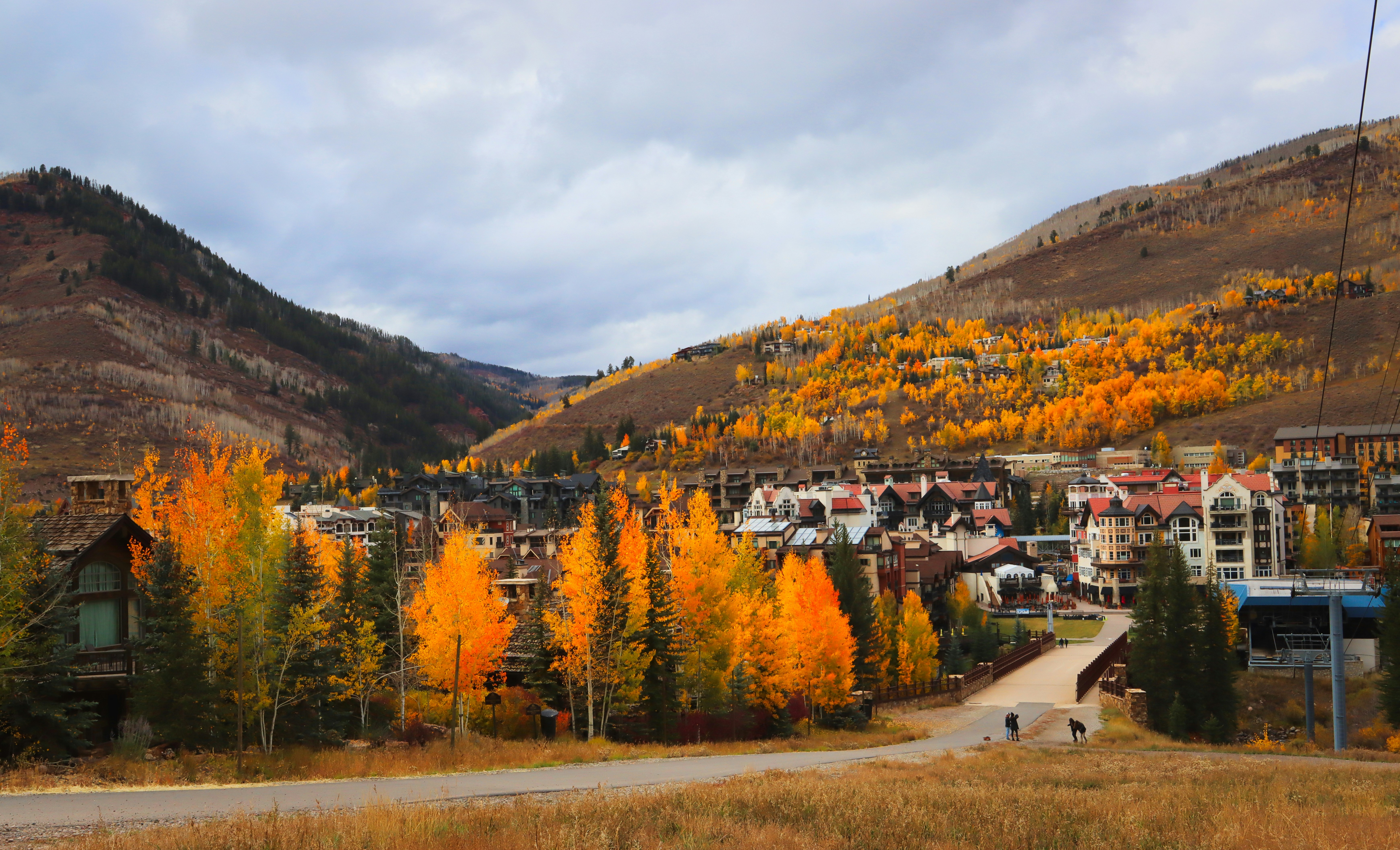 Vibrant aspen trees in golden hues surround a charming mountain village nestled among rolling hills. A serene path leads through the landscape.