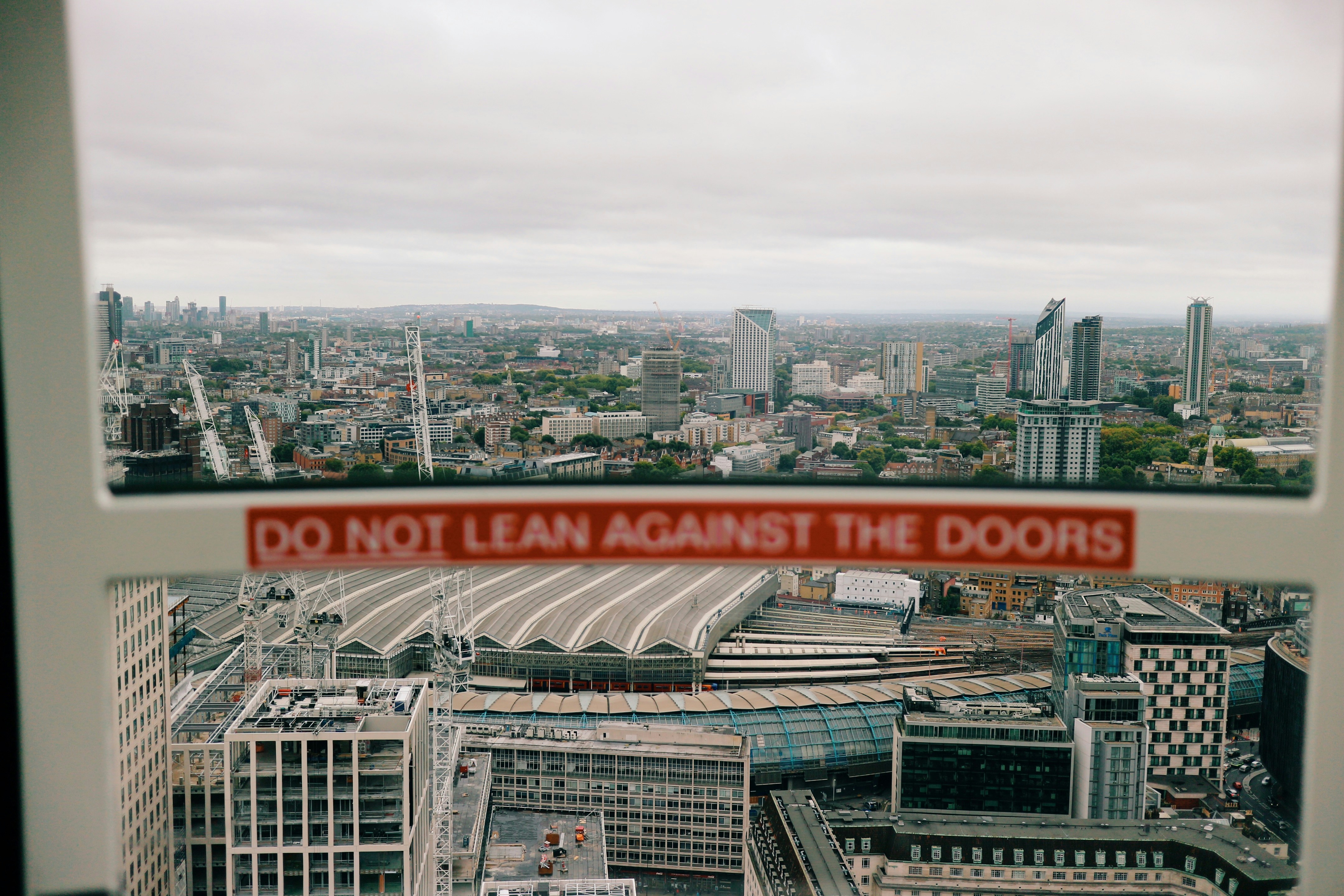 A panoramic view of a bustling cityscape framed by a safety warning on a glass panel. The scene captures a blend of modern architecture and urban life.