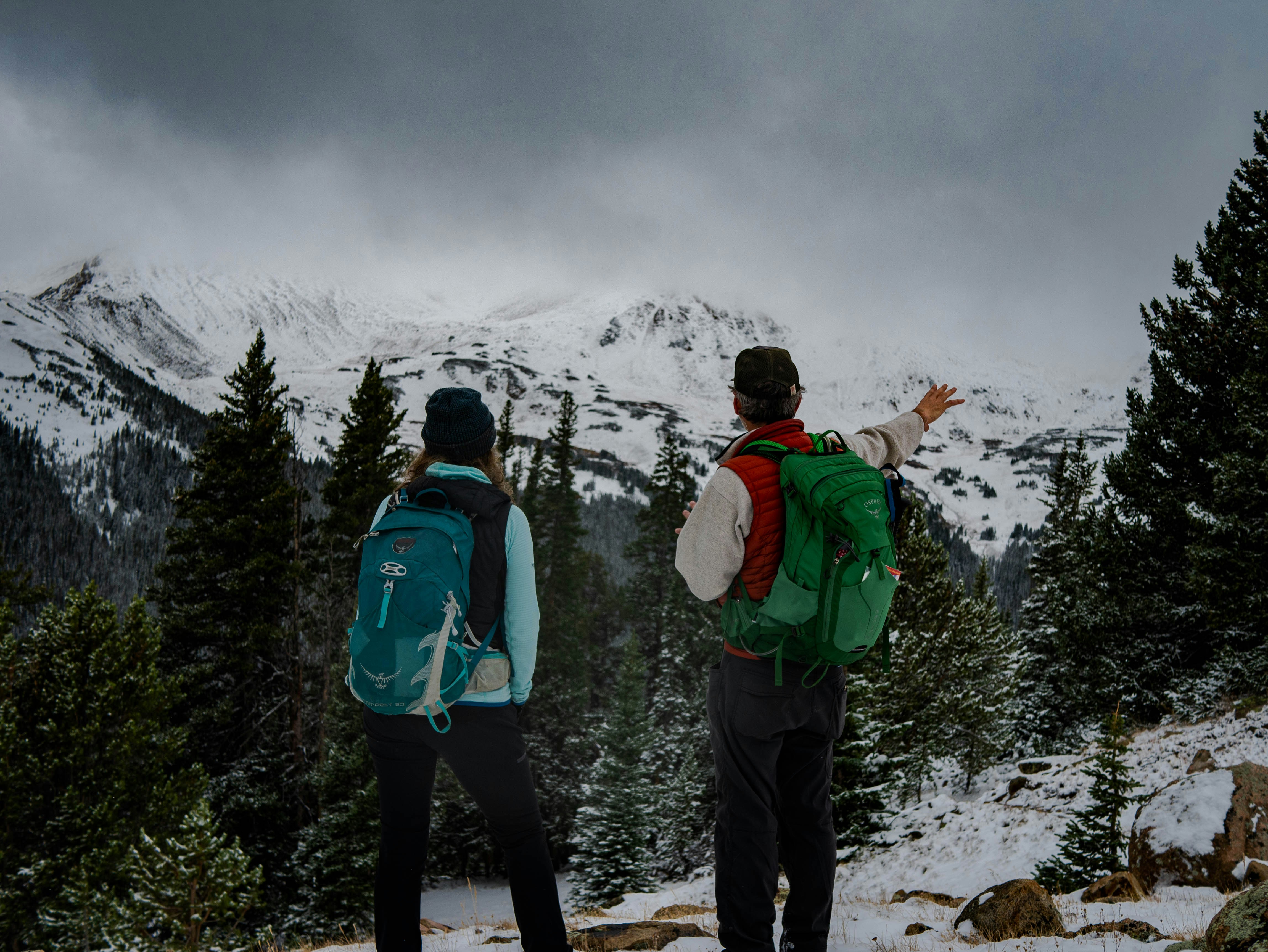 Two hikers stand amidst a snowy landscape, gazing at distant mountains while one gestures towards the peaks. Their backpacks suggest a journey into the wild.