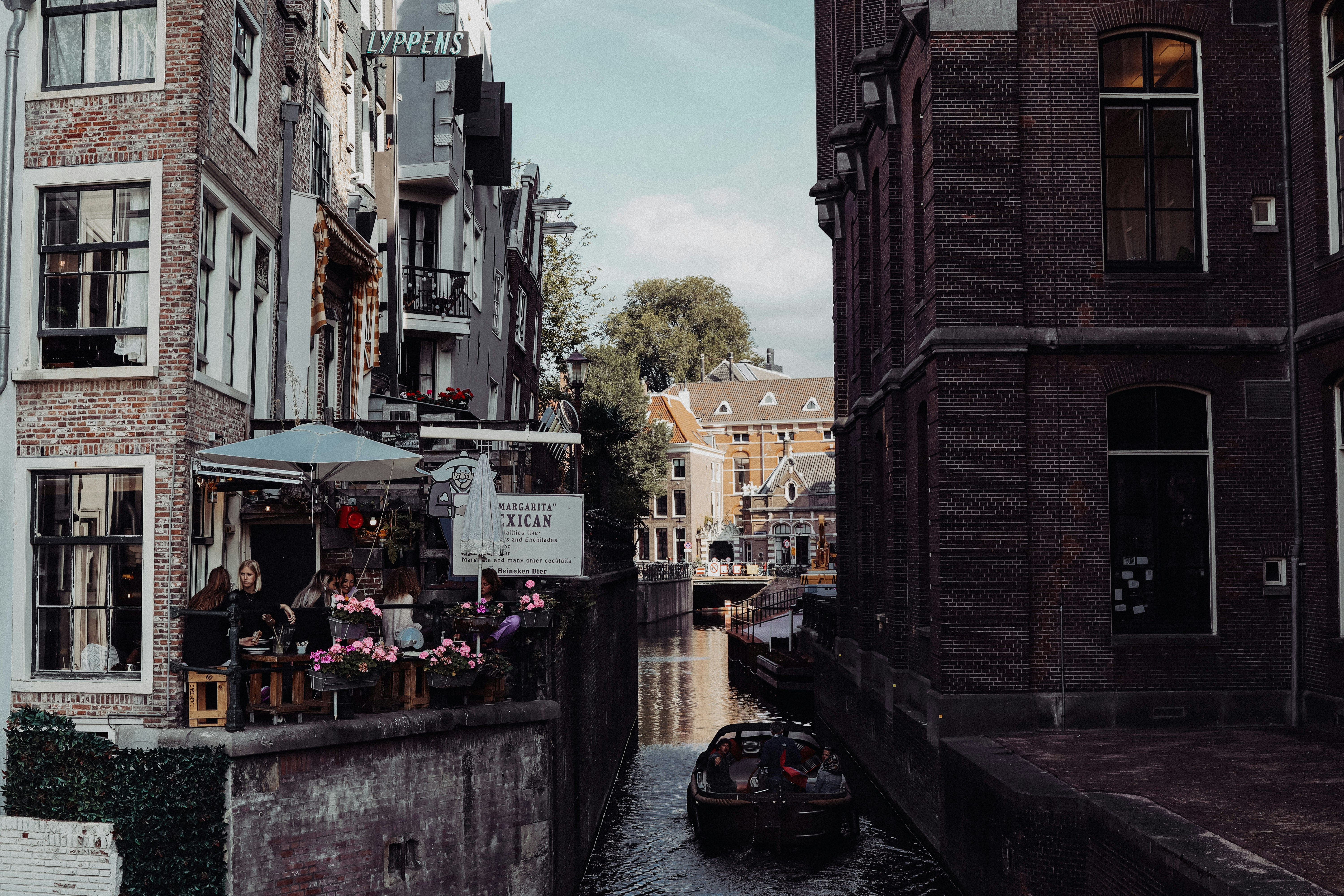 Charming canal-side café bustling with patrons amidst historic Dutch architecture. A small boat glides through the serene waters.