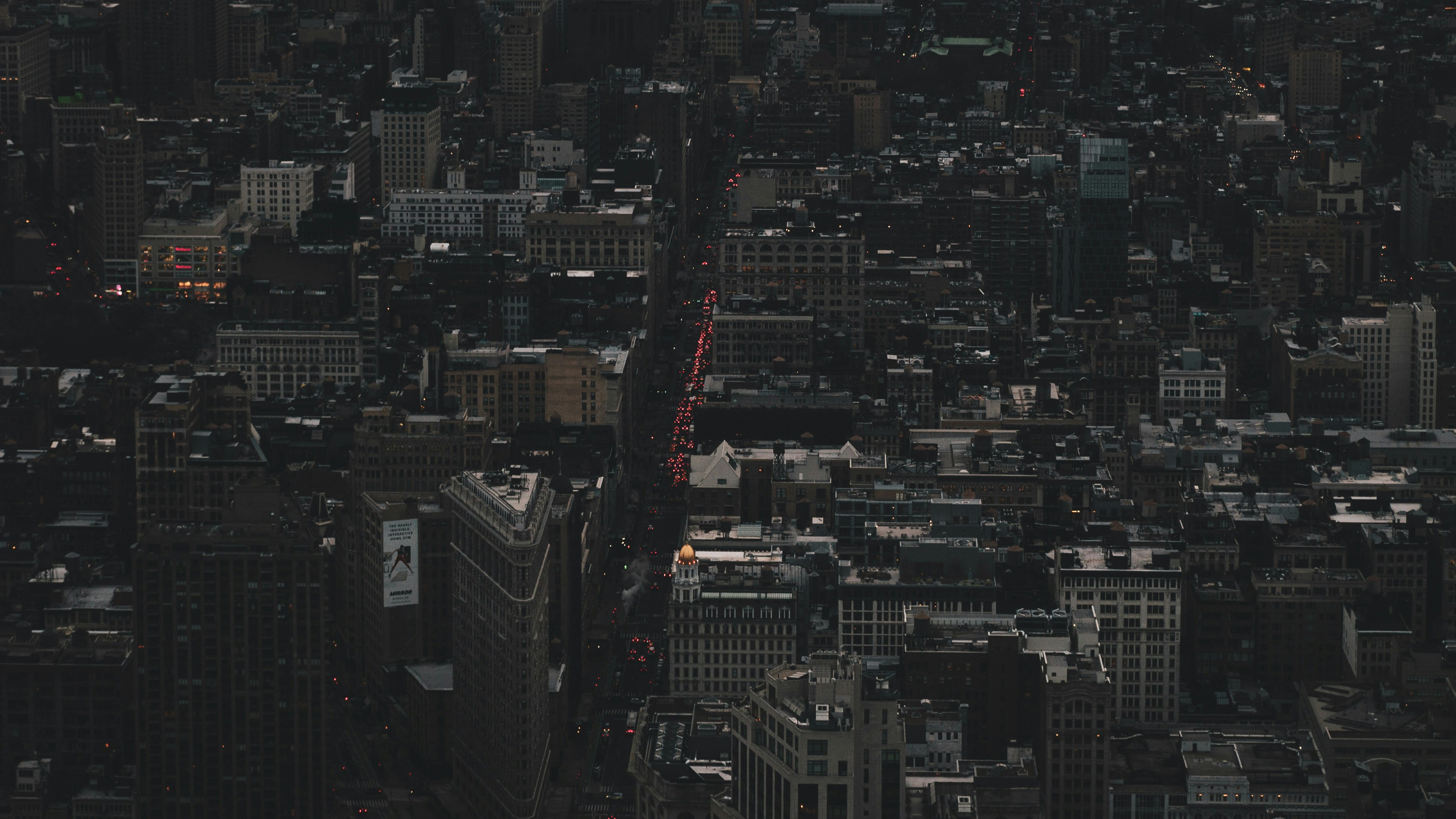 Aerial view of a bustling cityscape at dusk, illuminated by streaks of car lights along the main thoroughfare. The contrast of dark buildings against the glowing streets creates a dynamic urban atmosphere.