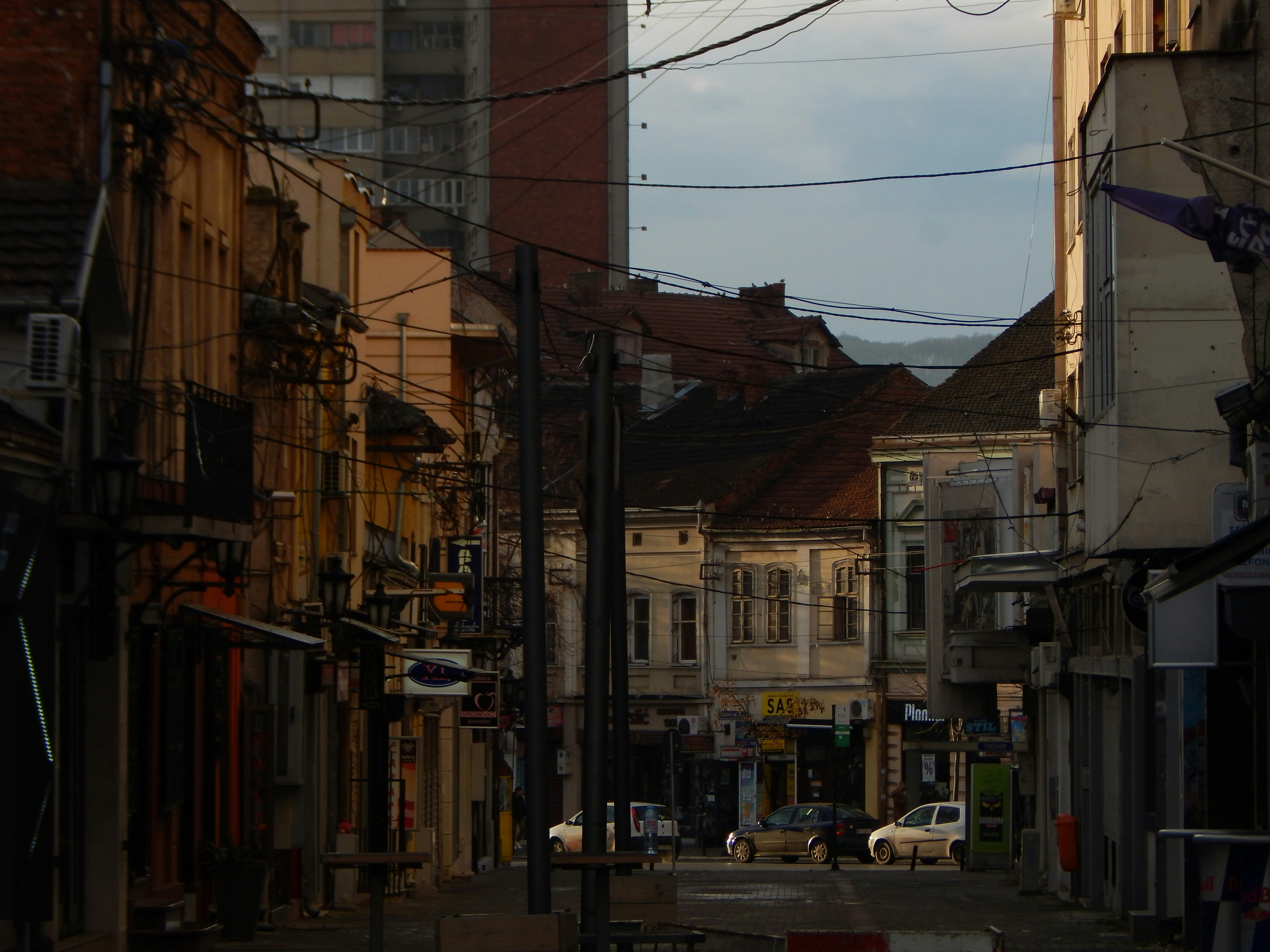 Narrow urban street lined with colorful facades and overhead wires extends toward a distant white car anchoring the scene.