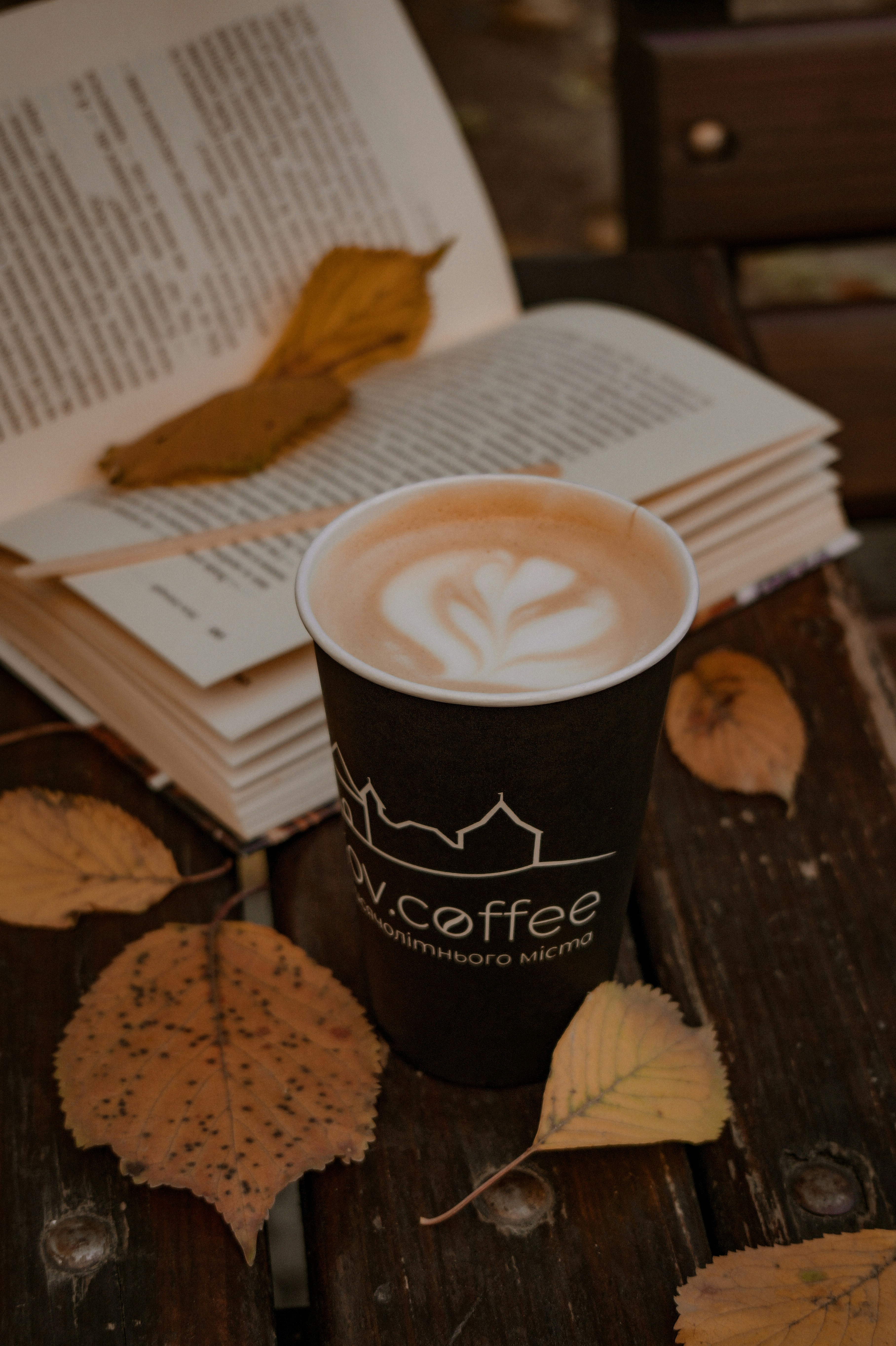 Photograph of a latte with latte art on a weathered wooden table, surrounded by autumn leaves and an open book. The scene emphasizes cozy study vibes.