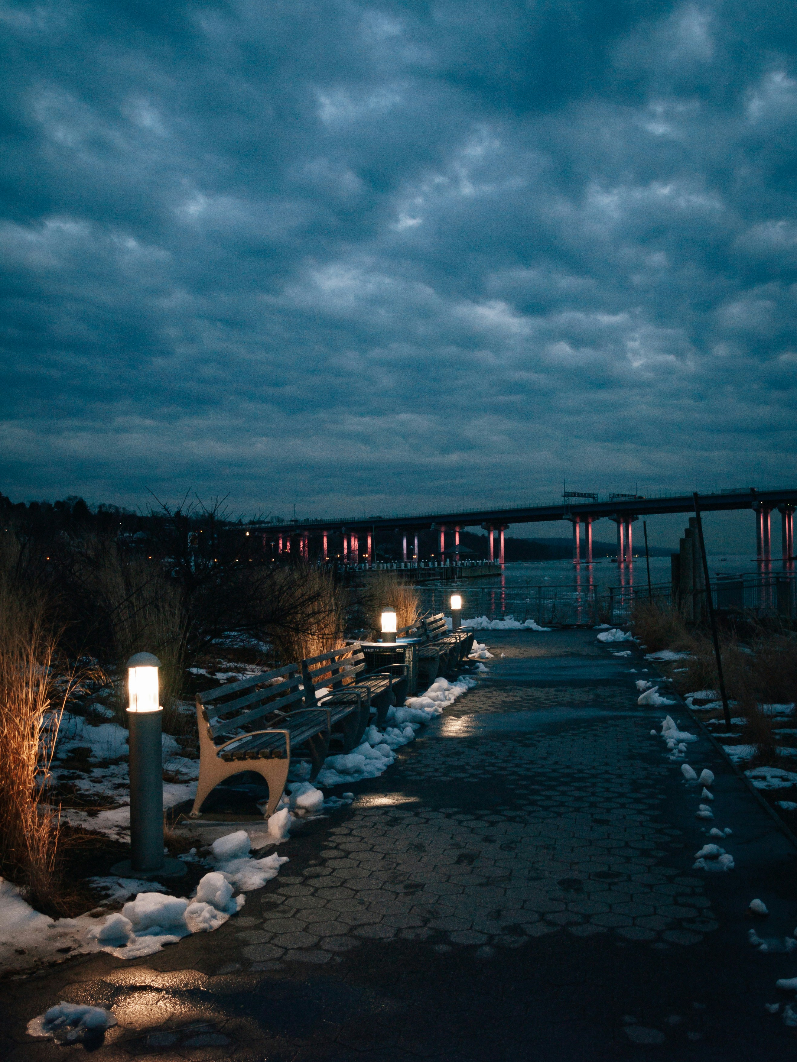 Photograph of a blue-hour coastal boardwalk with snow along the path, glowing lamps and benches, extending toward a distant red-lit bridge over the water.