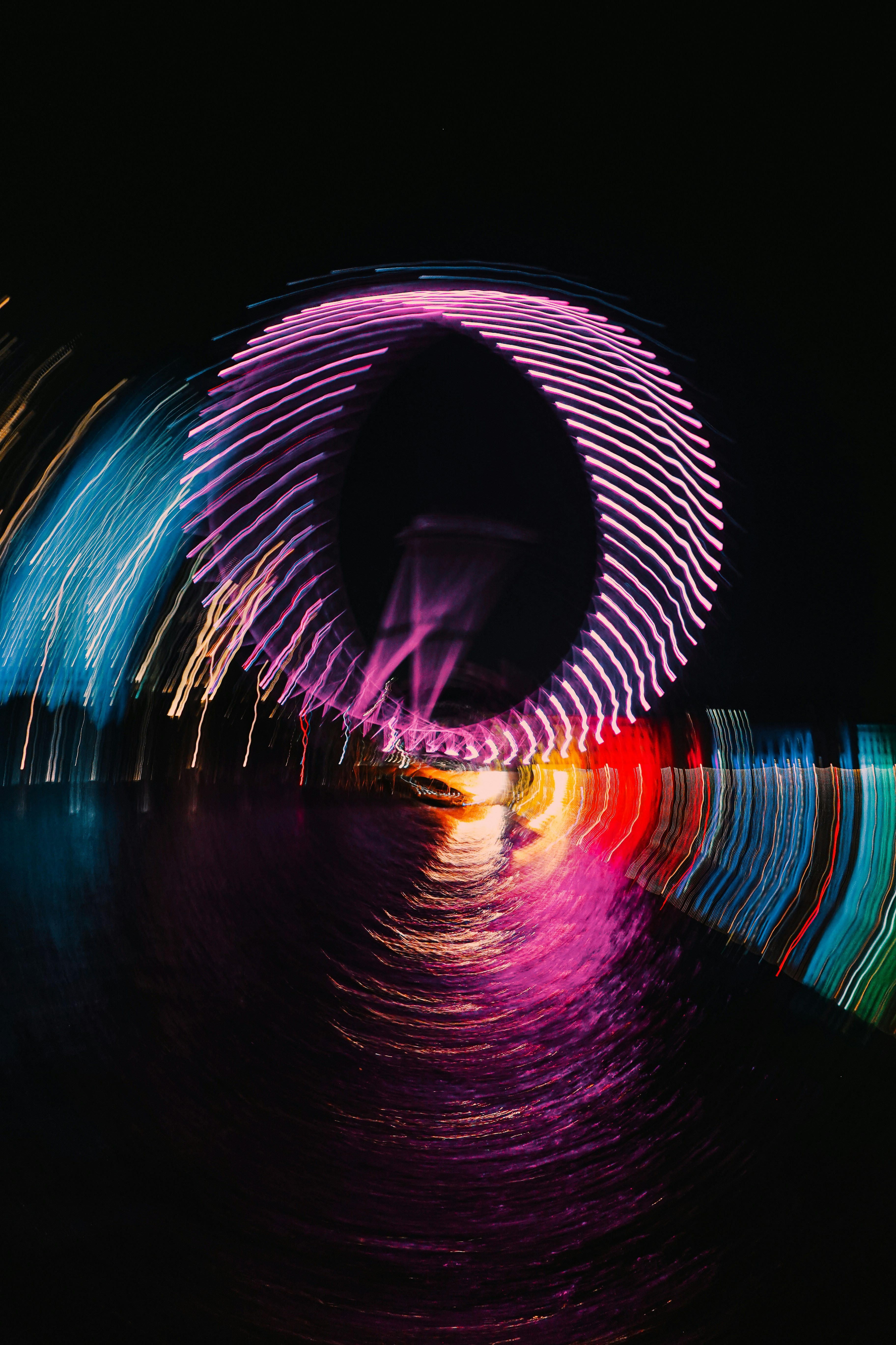 Long-exposure shot of a Ferris wheel's neon ring spinning against the night sky, with vivid pink and purple arcs and reflections on the water.